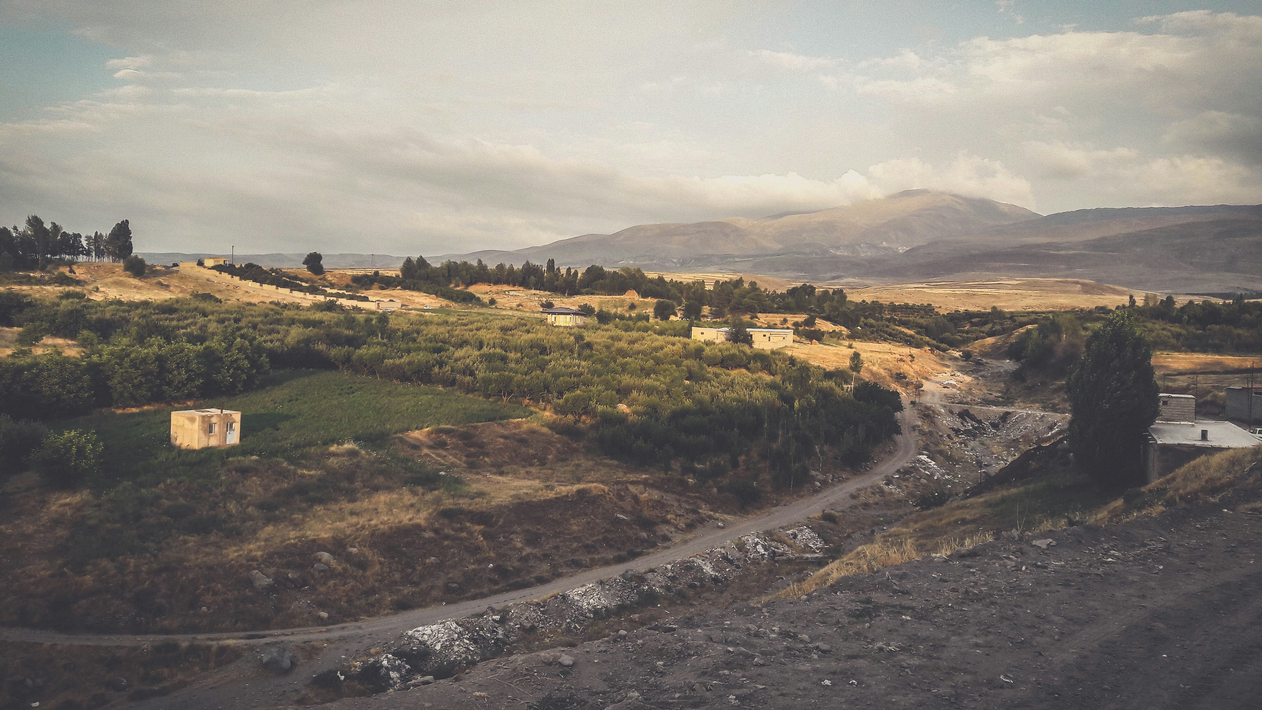 Landscape photograph of rolling hills with scattered square buildings and a winding dirt road under a cloudy sky.