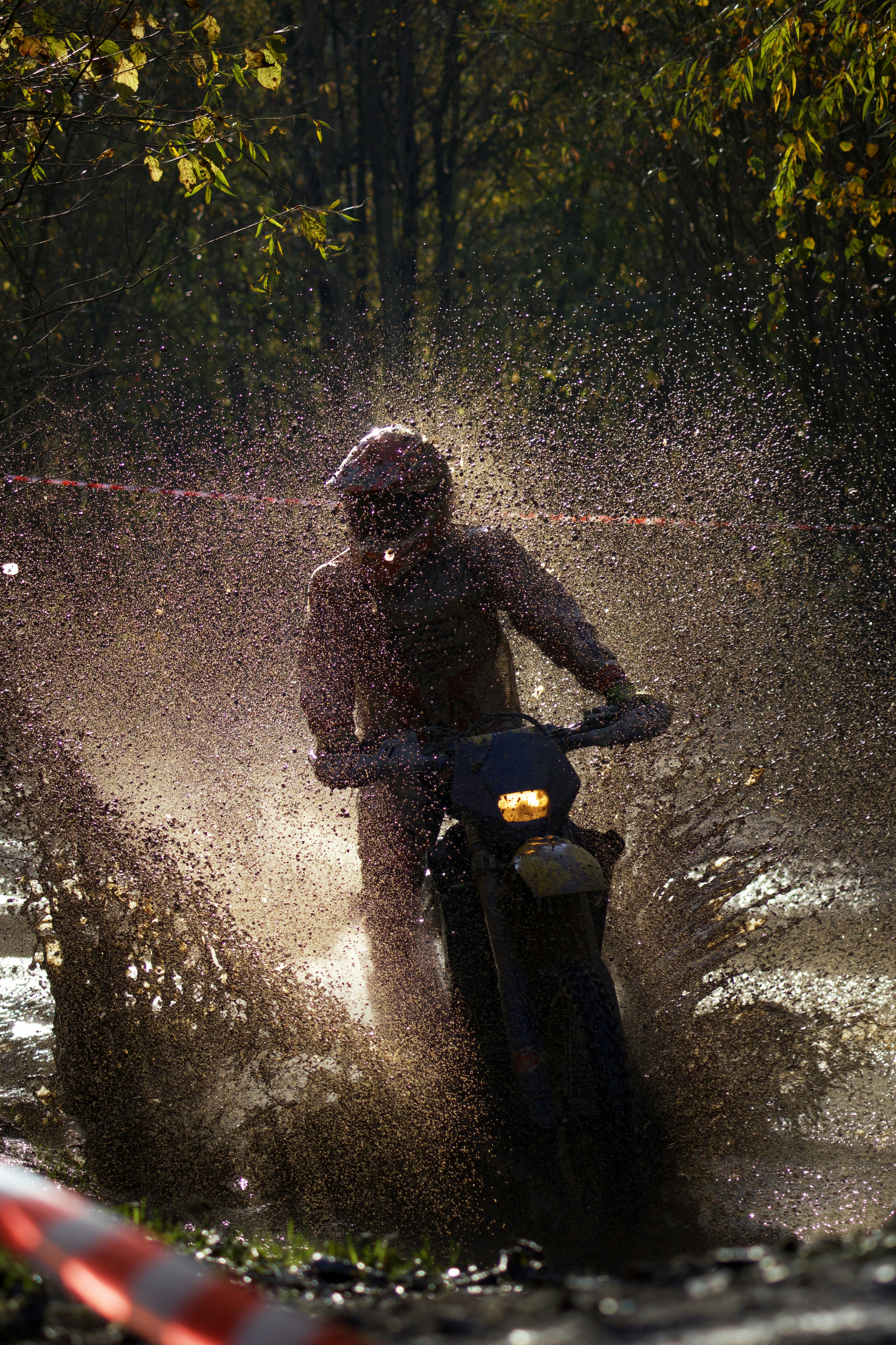 Motorcycle rider on a dirt bike splashes through a forest puddle, sending a curtain of spray around a dark silhouette as sunlight filters through leaves.