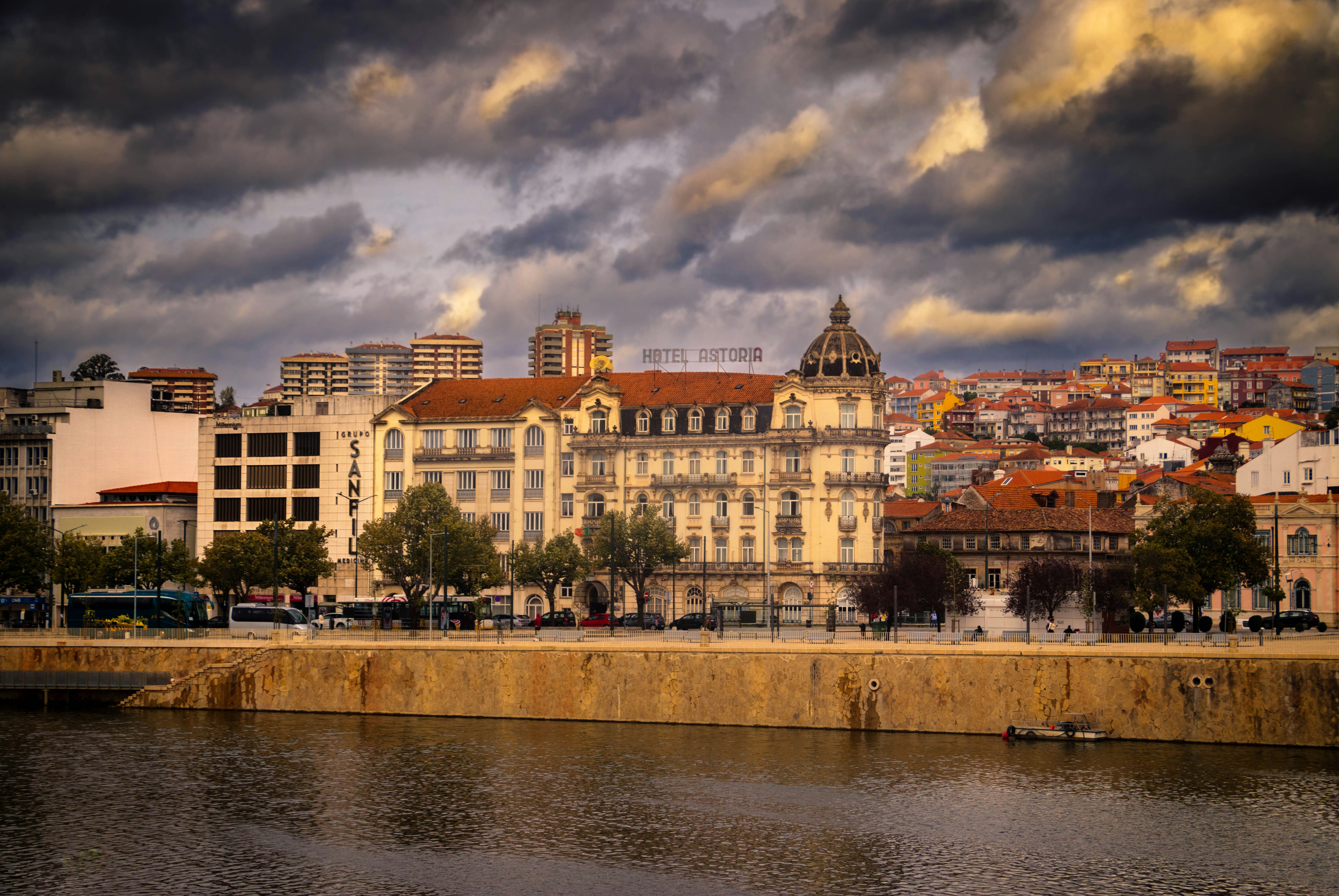 Historic buildings line the riverbank under a dramatic, cloud-filled sky at sunset.