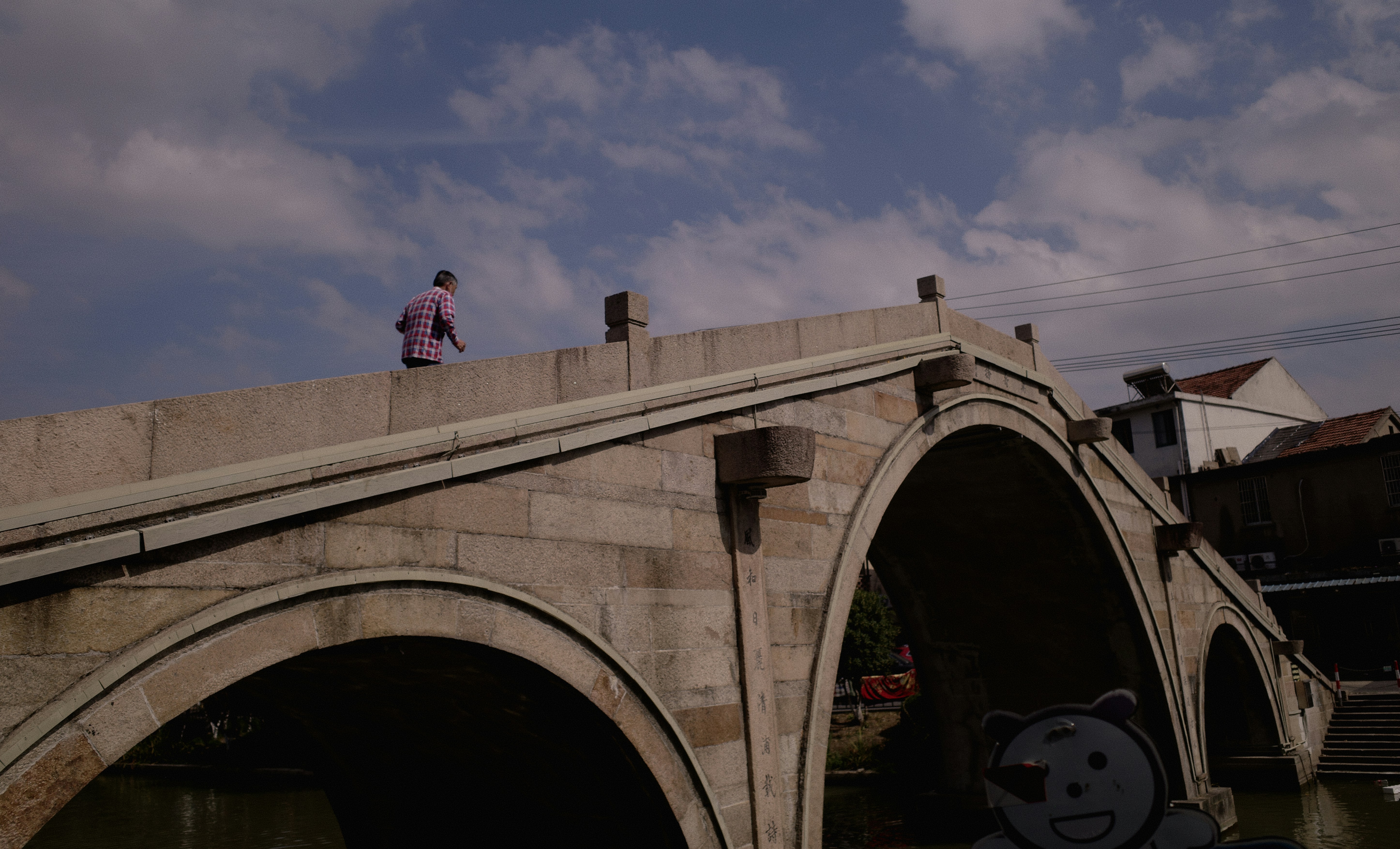 Stone-arch bridge over a canal under a blue sky; a lone figure stands atop the parapet on the left, and a round cartoon logo sits at the lower-right.