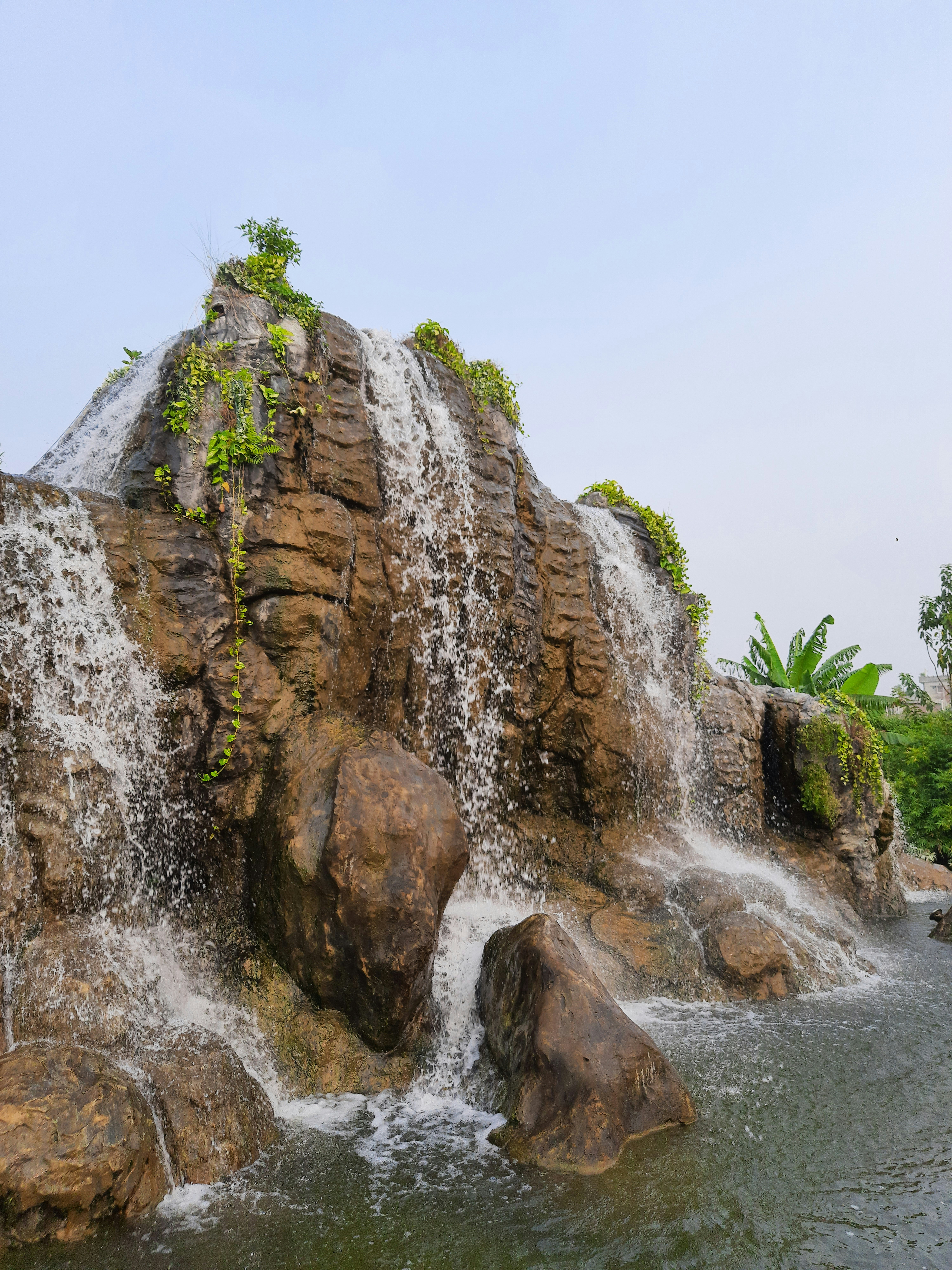 Rocky cliff with cascading water and green vines over a calm pool under a clear blue sky.
