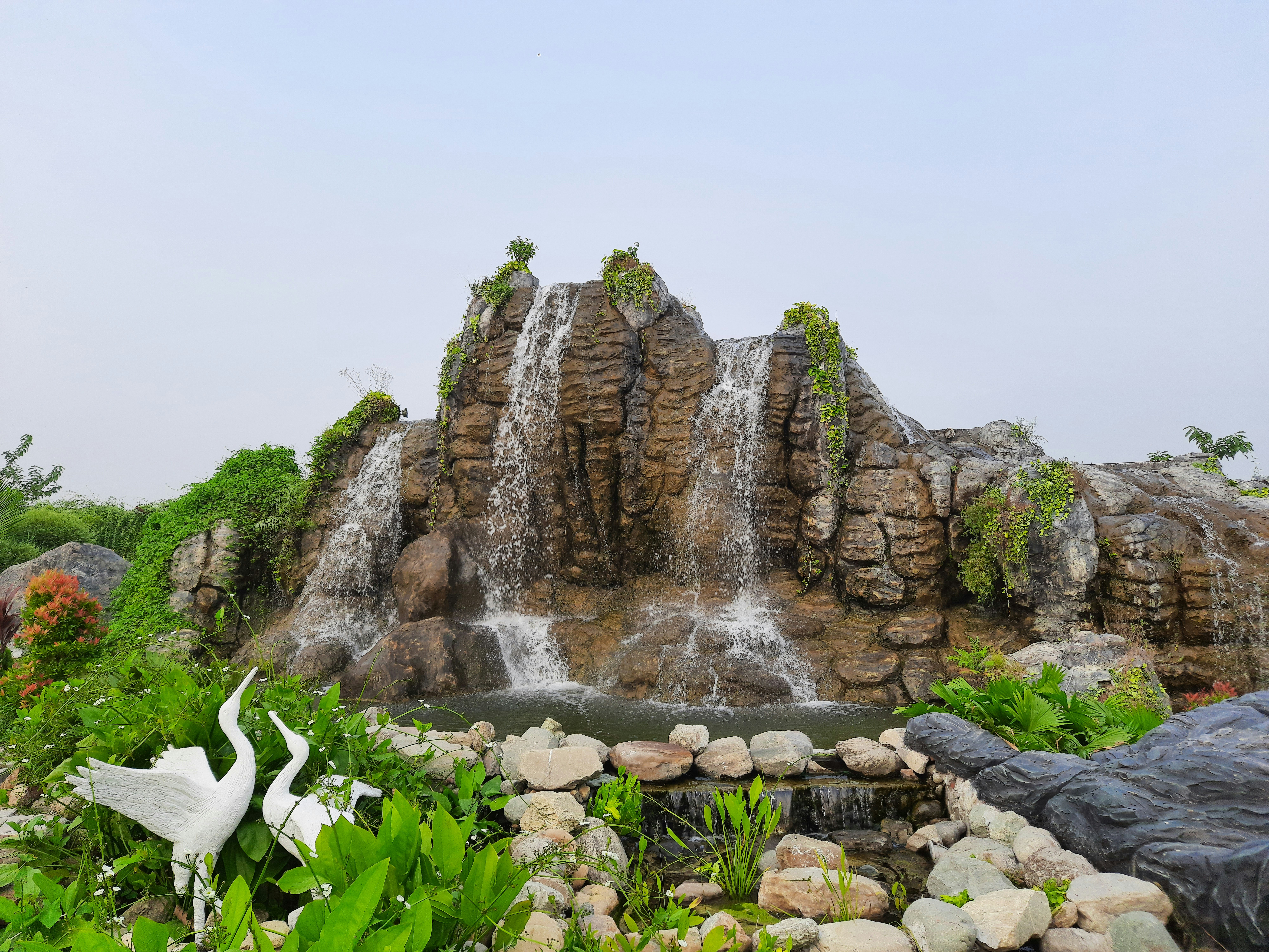A landscape photograph of a rock waterfall garden with lush vegetation, a tranquil pool, and a white crane sculpture in the foreground.