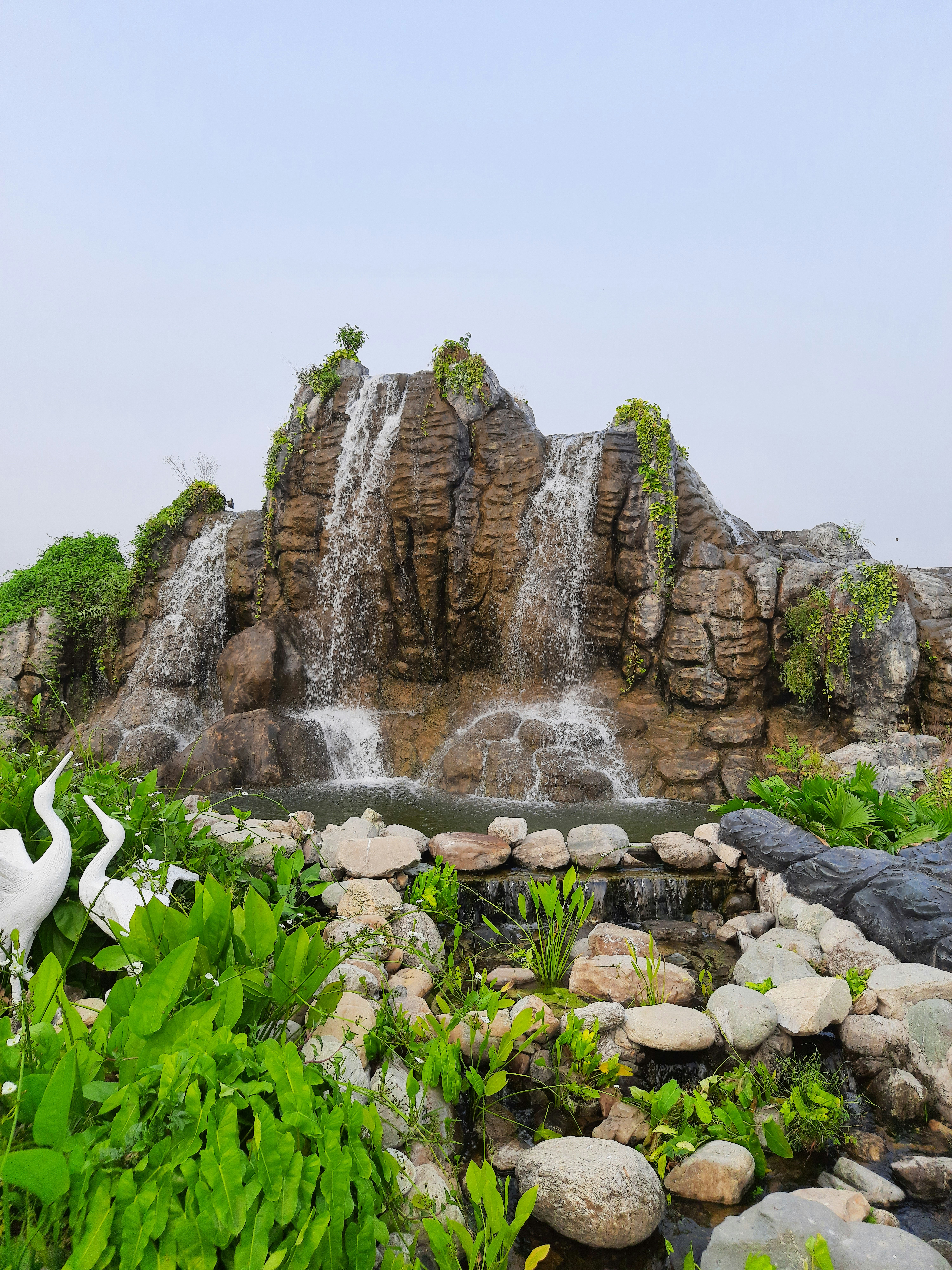 A tranquil waterfall flows over rocky formations, surrounded by lush greenery and decorative stones. Two elegant white cranes stand gracefully in the foreground.
