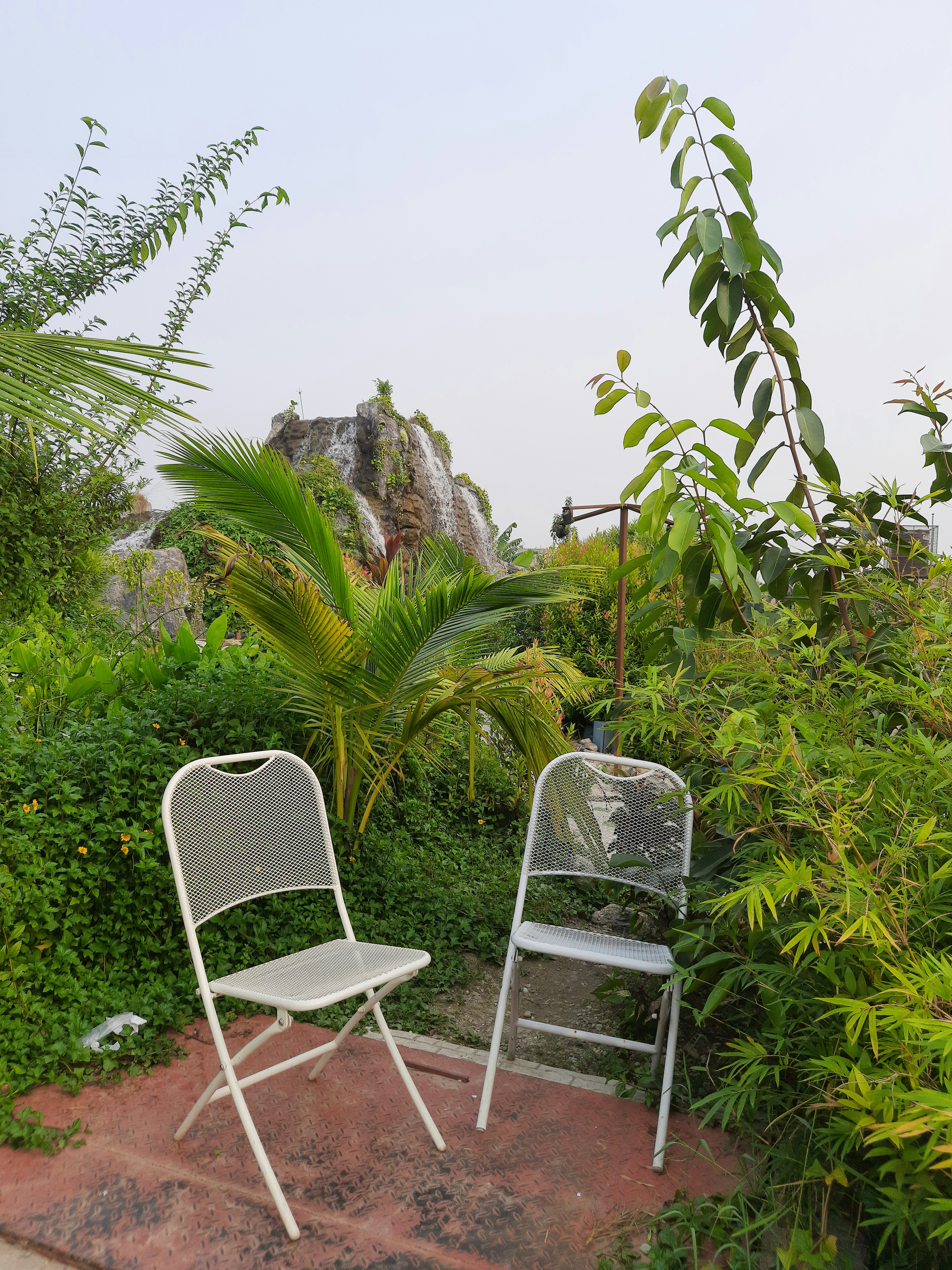 Two white metal chairs sit on a red-tiled patio, surrounded by dense tropical plants and a rocky outcrop in the background.
