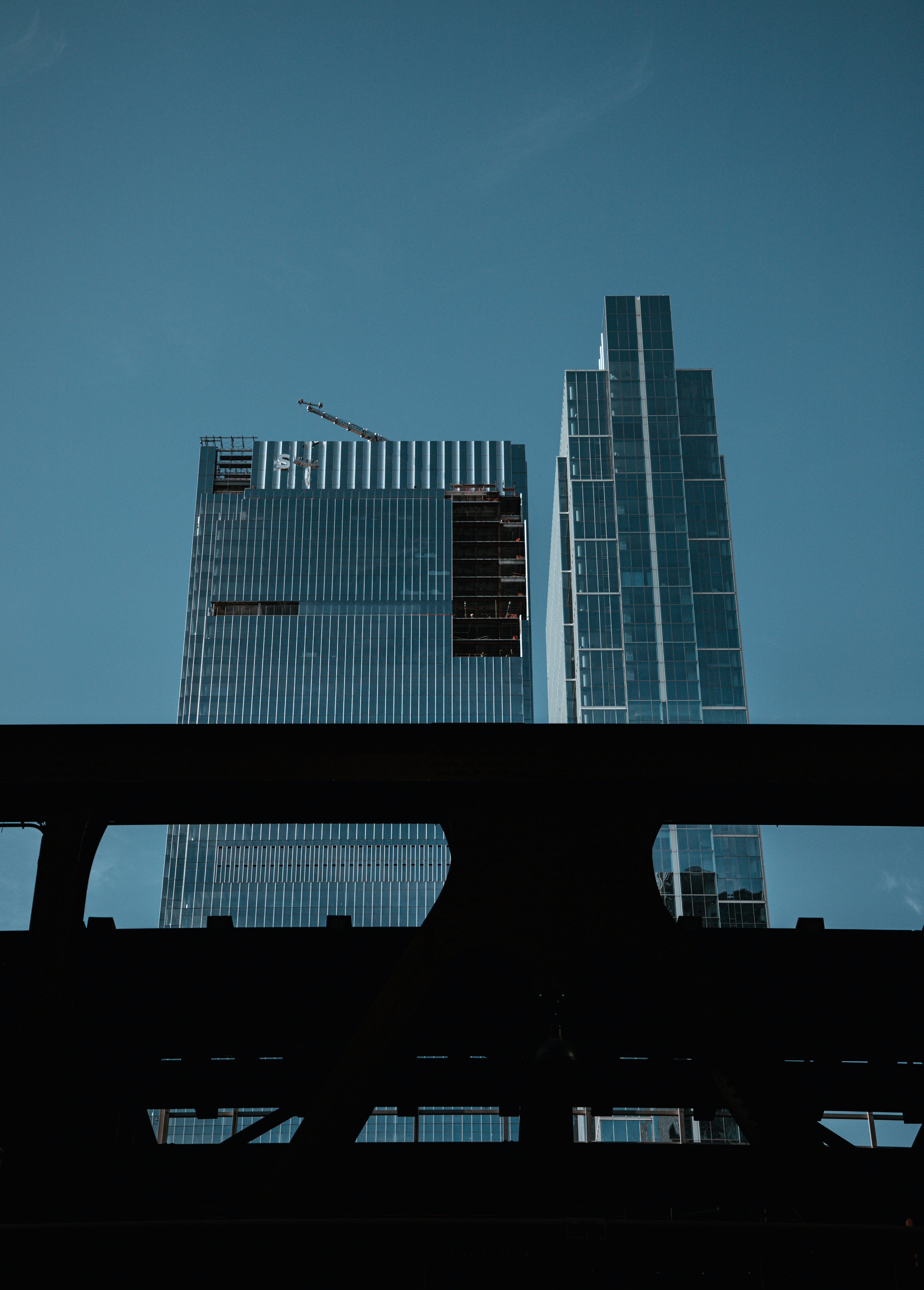 Two towering skyscrapers rise against a clear blue sky, framed by the silhouette of an industrial structure below.