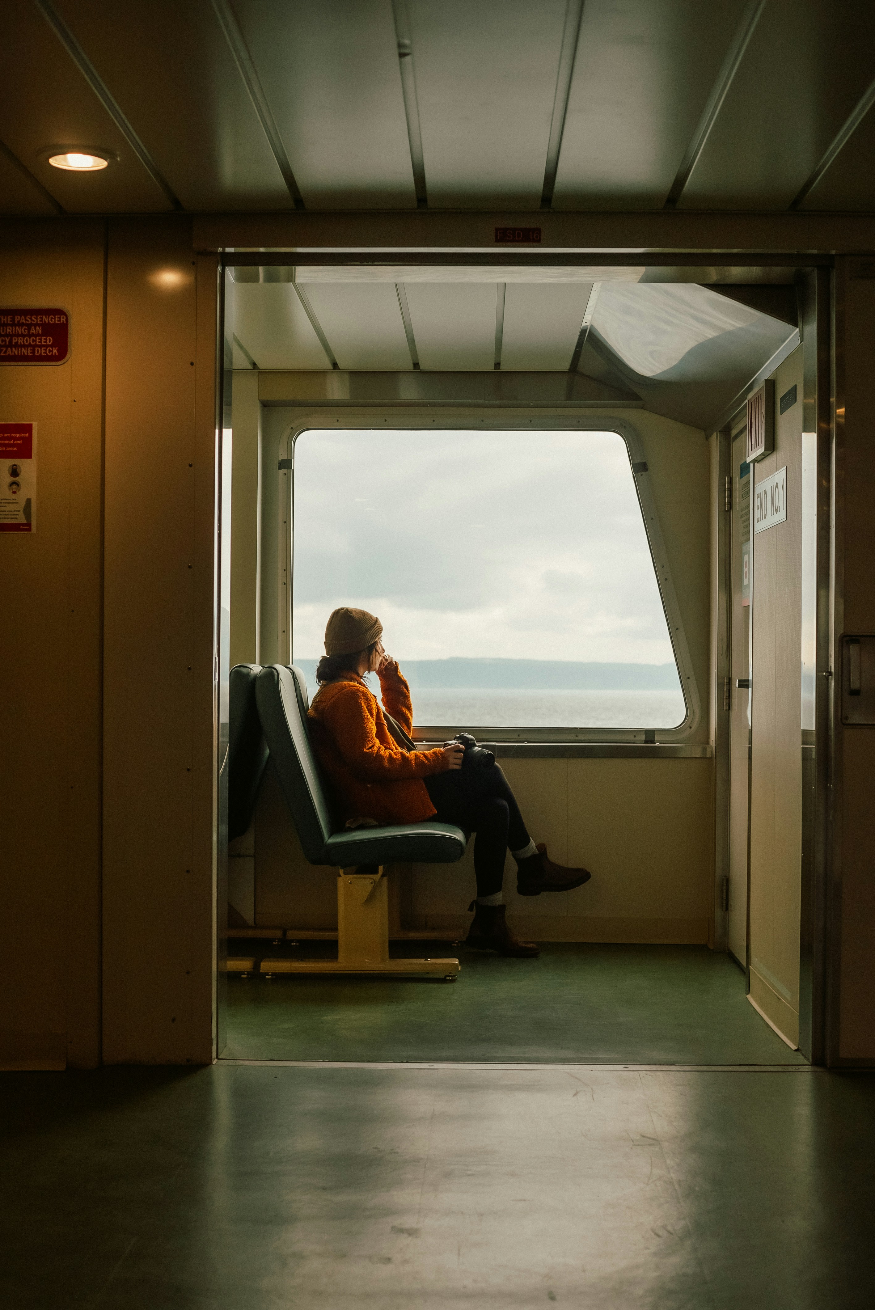 A person in an orange sweater sits contemplatively by a window on a ferry, gazing at the serene seascape beyond. The scene captures a blend of interior and exterior worlds.