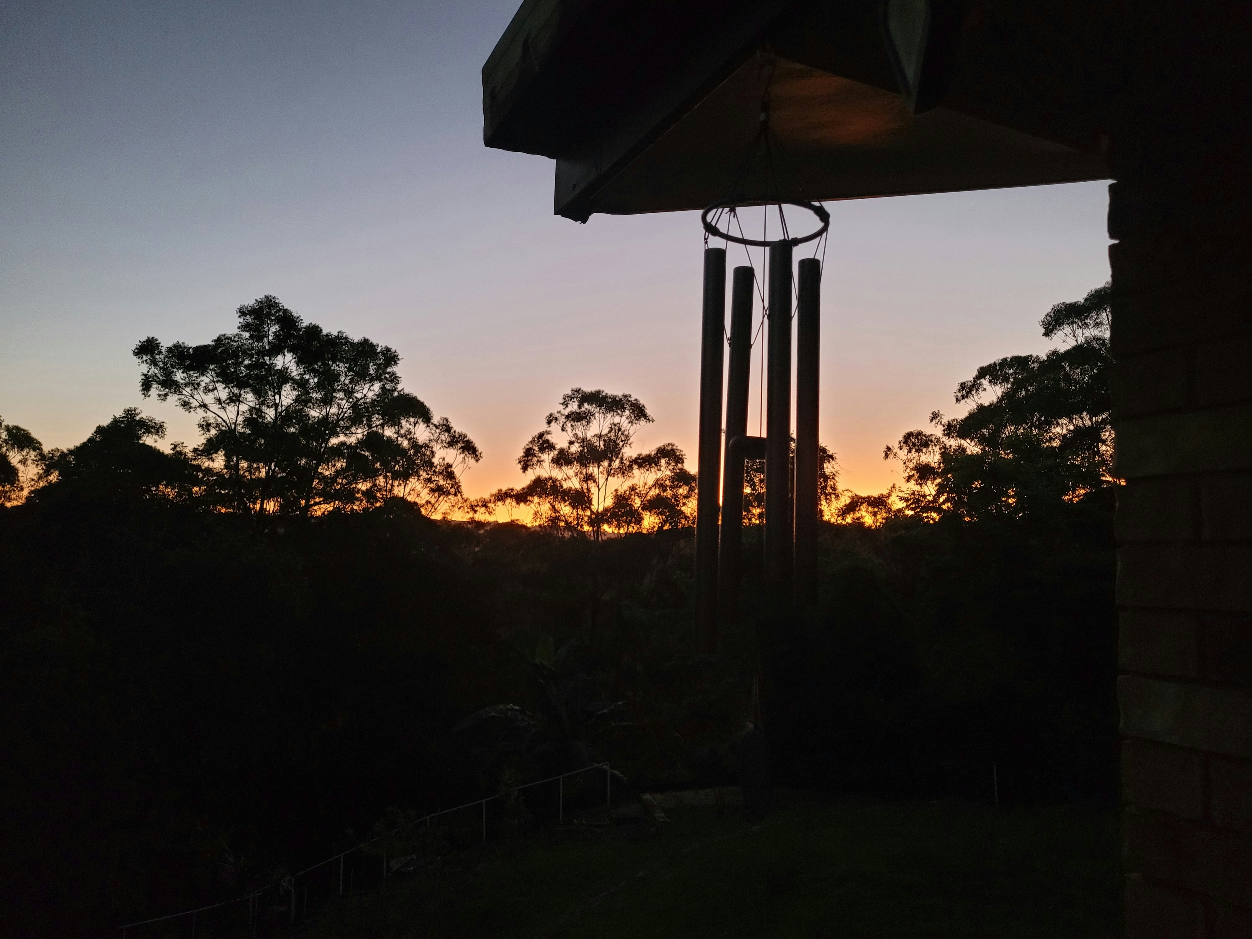 Wind chimes gently swaying against a backdrop of vibrant twilight hues and silhouetted trees. The scene captures the tranquil transition from day to night.