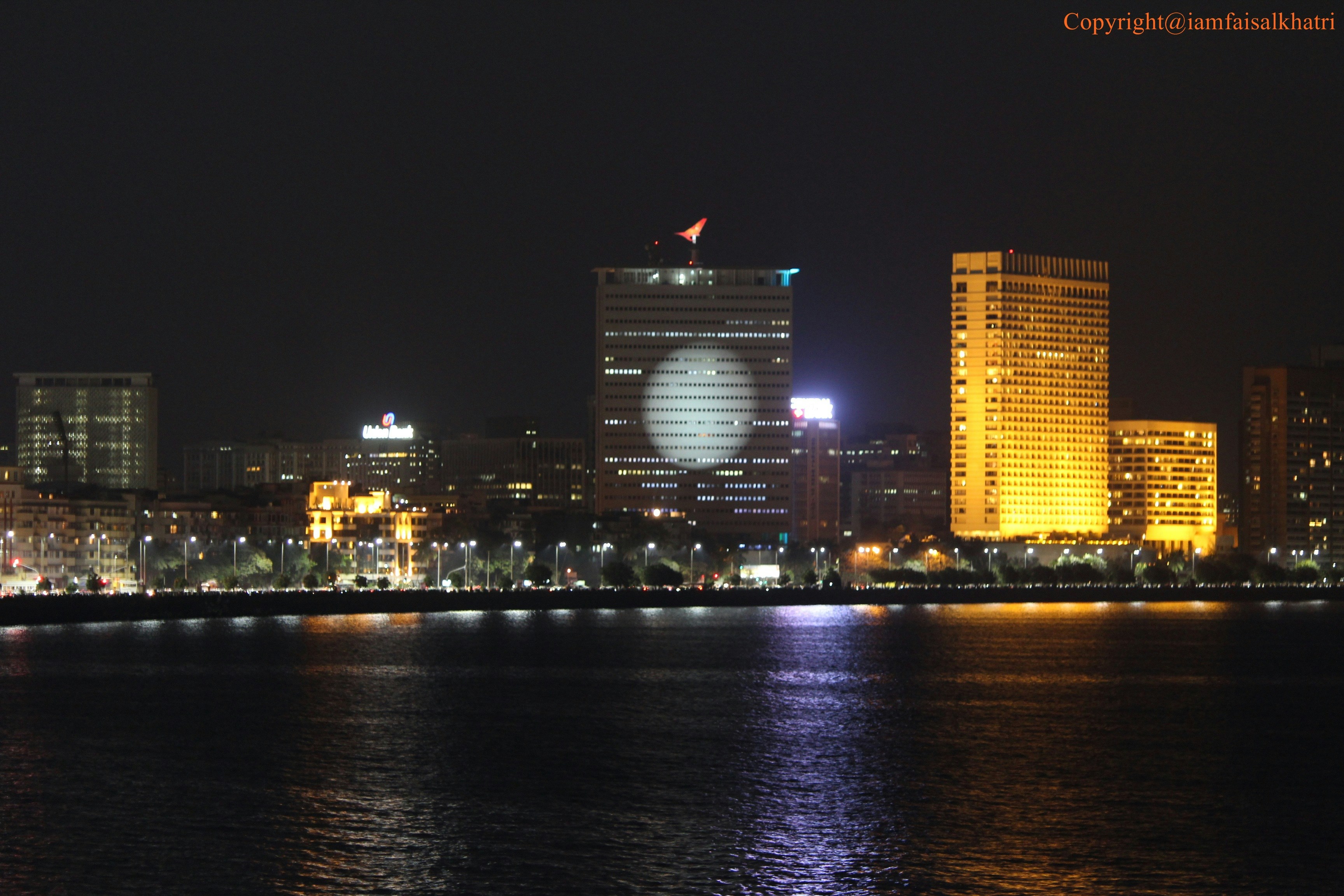 City skyline illuminated at night, featuring prominent buildings reflecting on the water's surface.