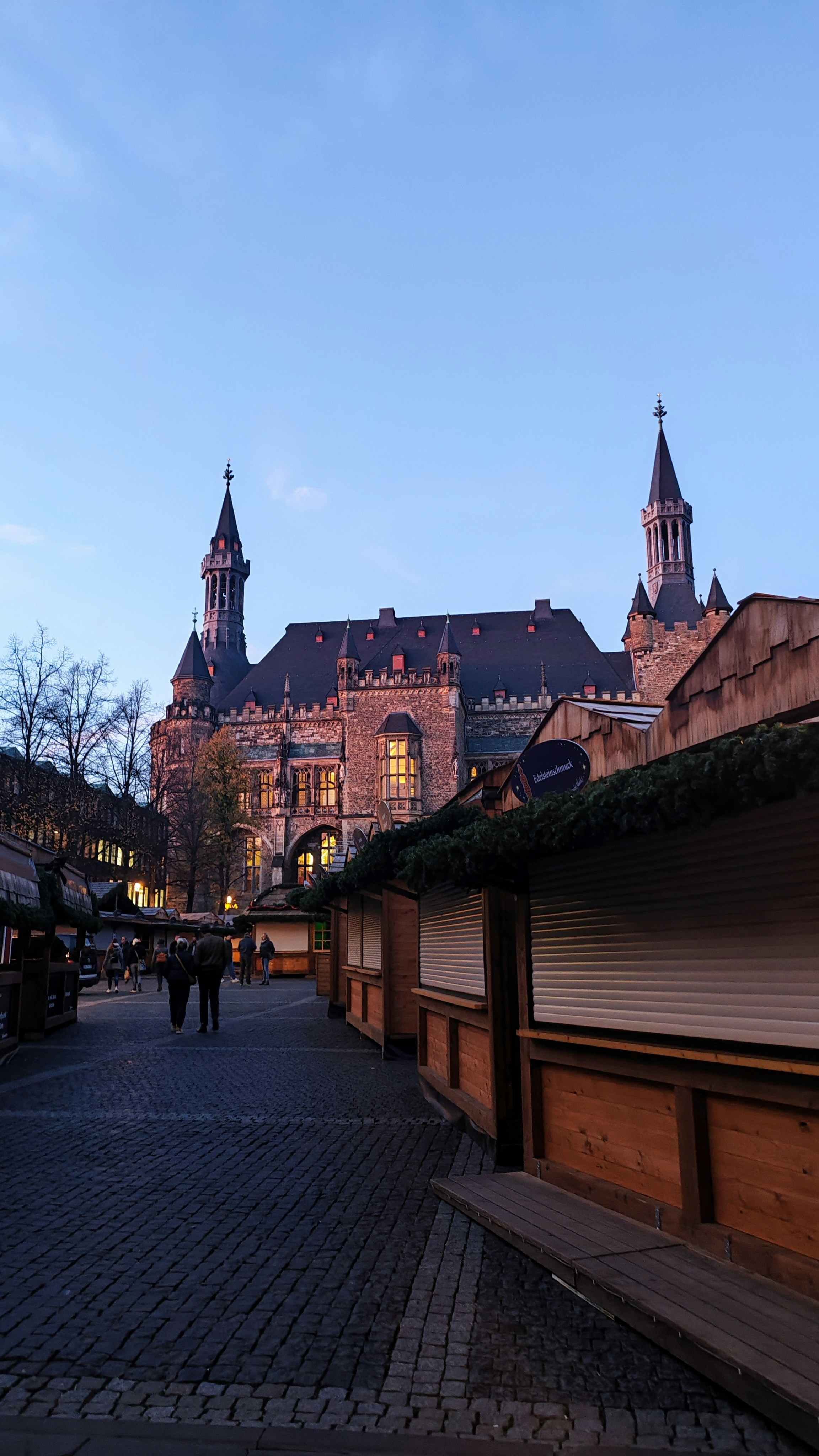Blue hour street scene showing a Gothic town hall with twin towers along a cobblestone market street and wooden stalls. The shot emphasizes architectural detail and atmospheric dusk lighting.