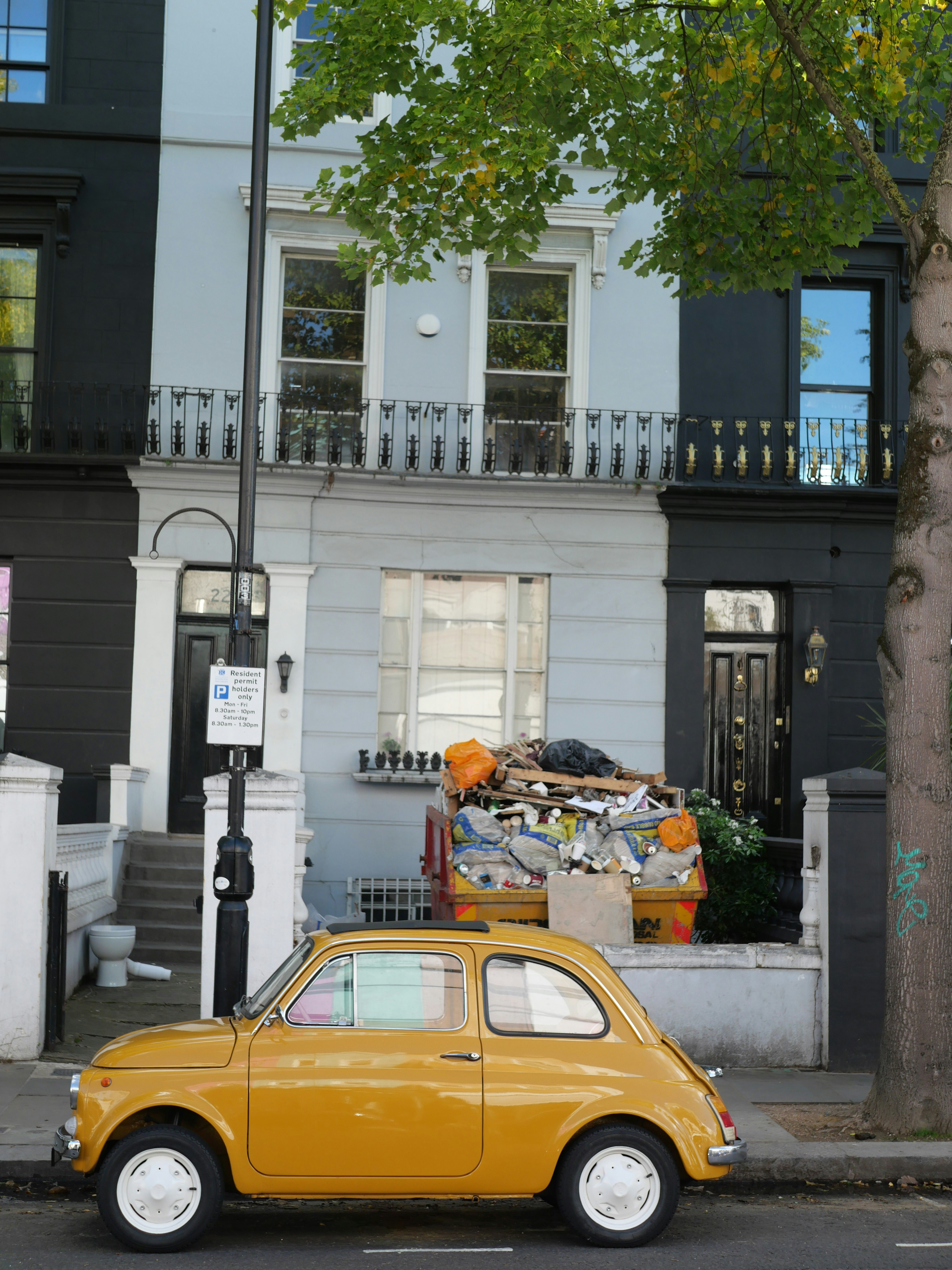 A classic yellow car parked beside a modern building, with a dumpster overflowing with debris in the background.