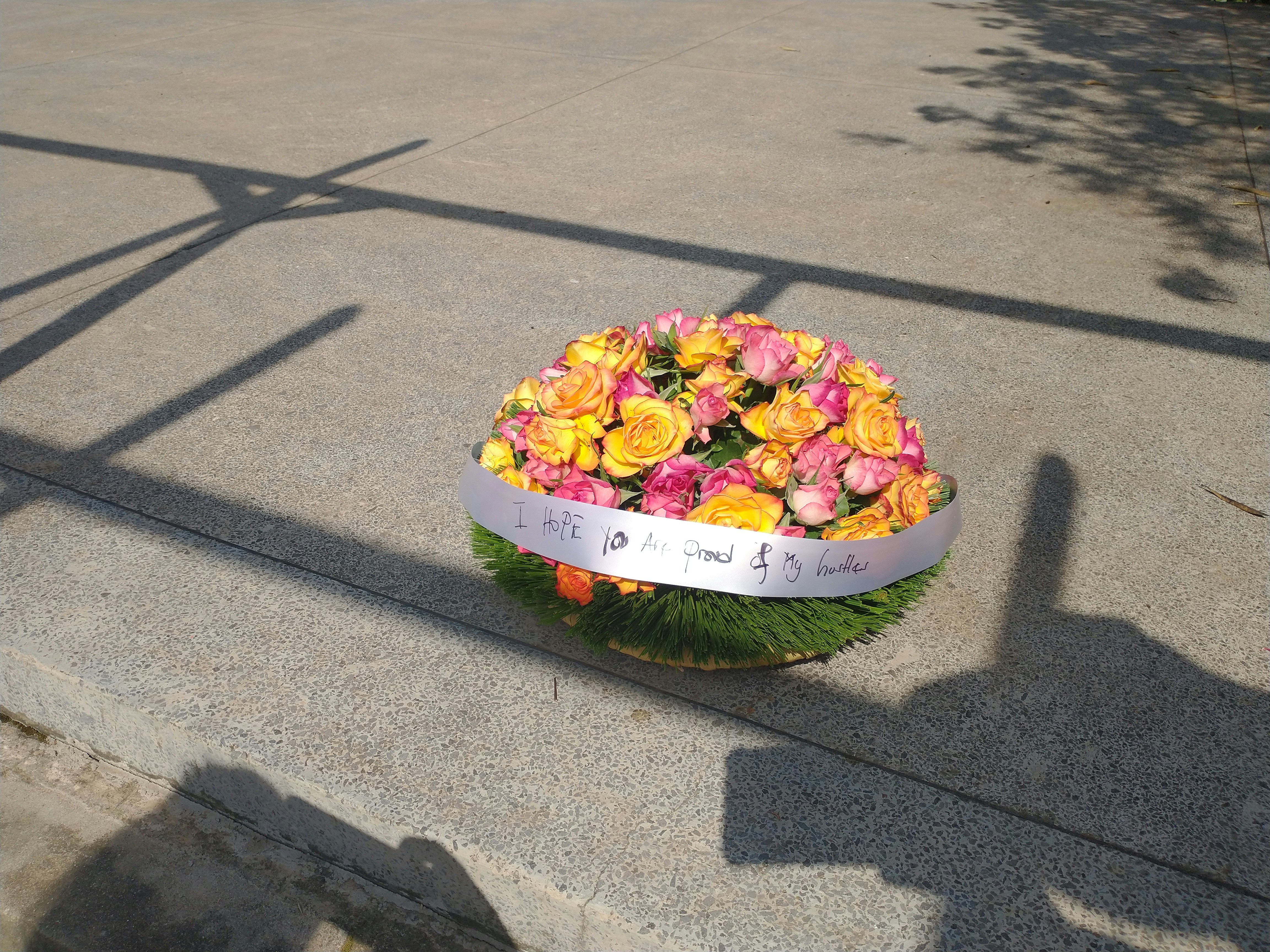 Wreath of colorful flowers with a commemorative ribbon on a sunlit pavement.