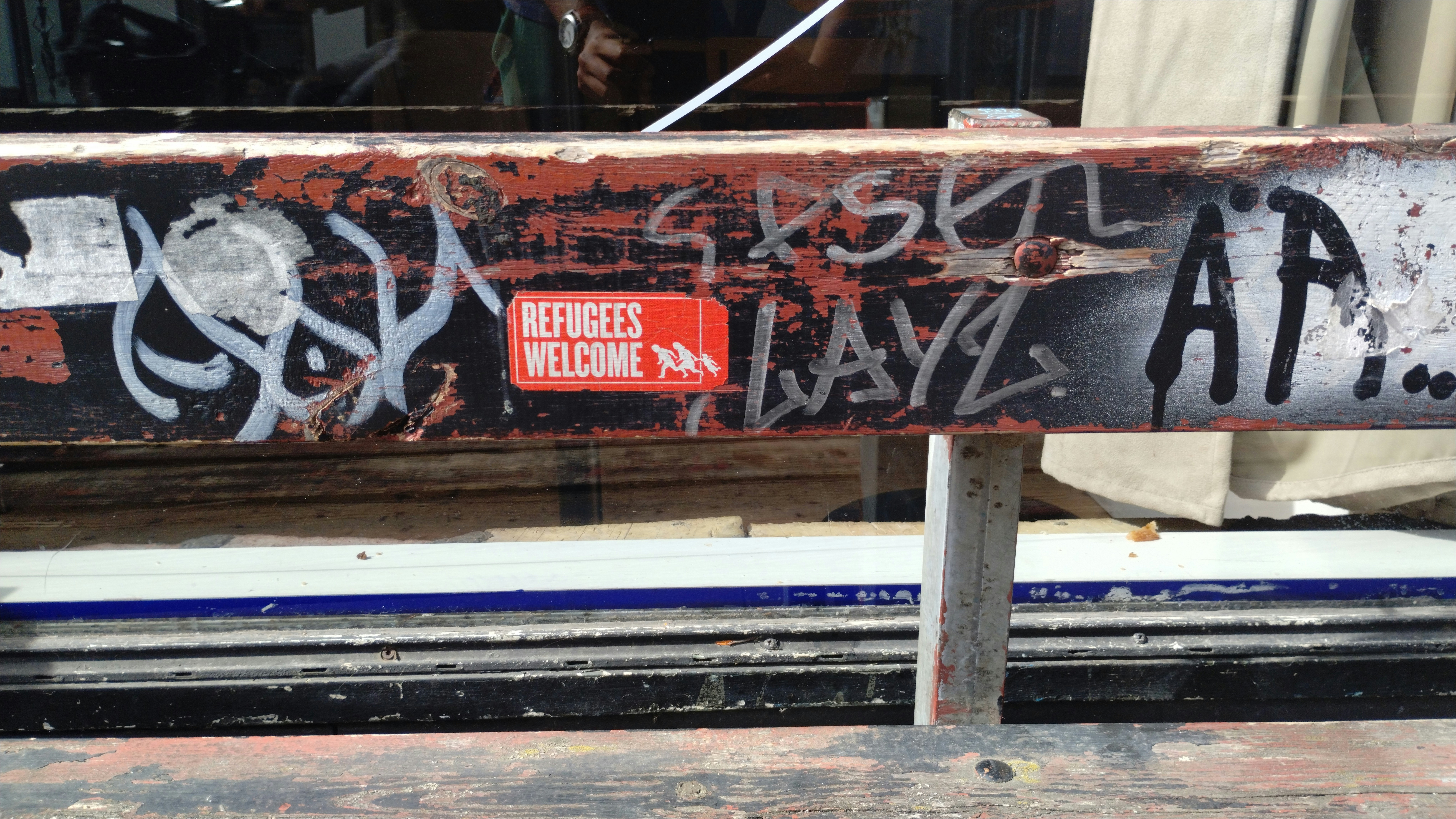 Graffiti and a bold sticker proclaiming 'Refugees Welcome' on a worn wooden surface, reflecting urban culture and social messages.