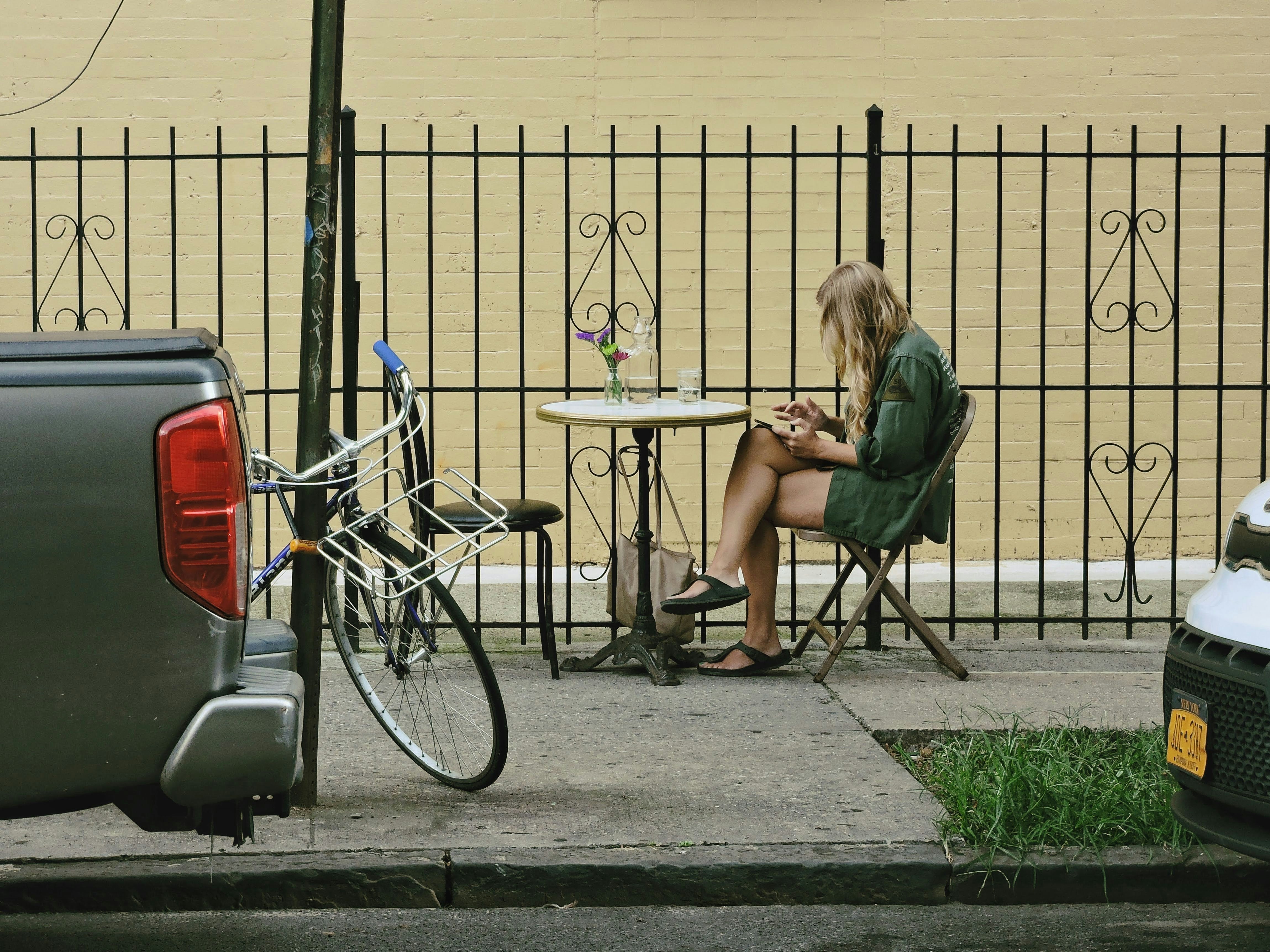 A young woman engrossed in her phone sits alone at a small table outside, surrounded by a bicycle and parked vehicles.