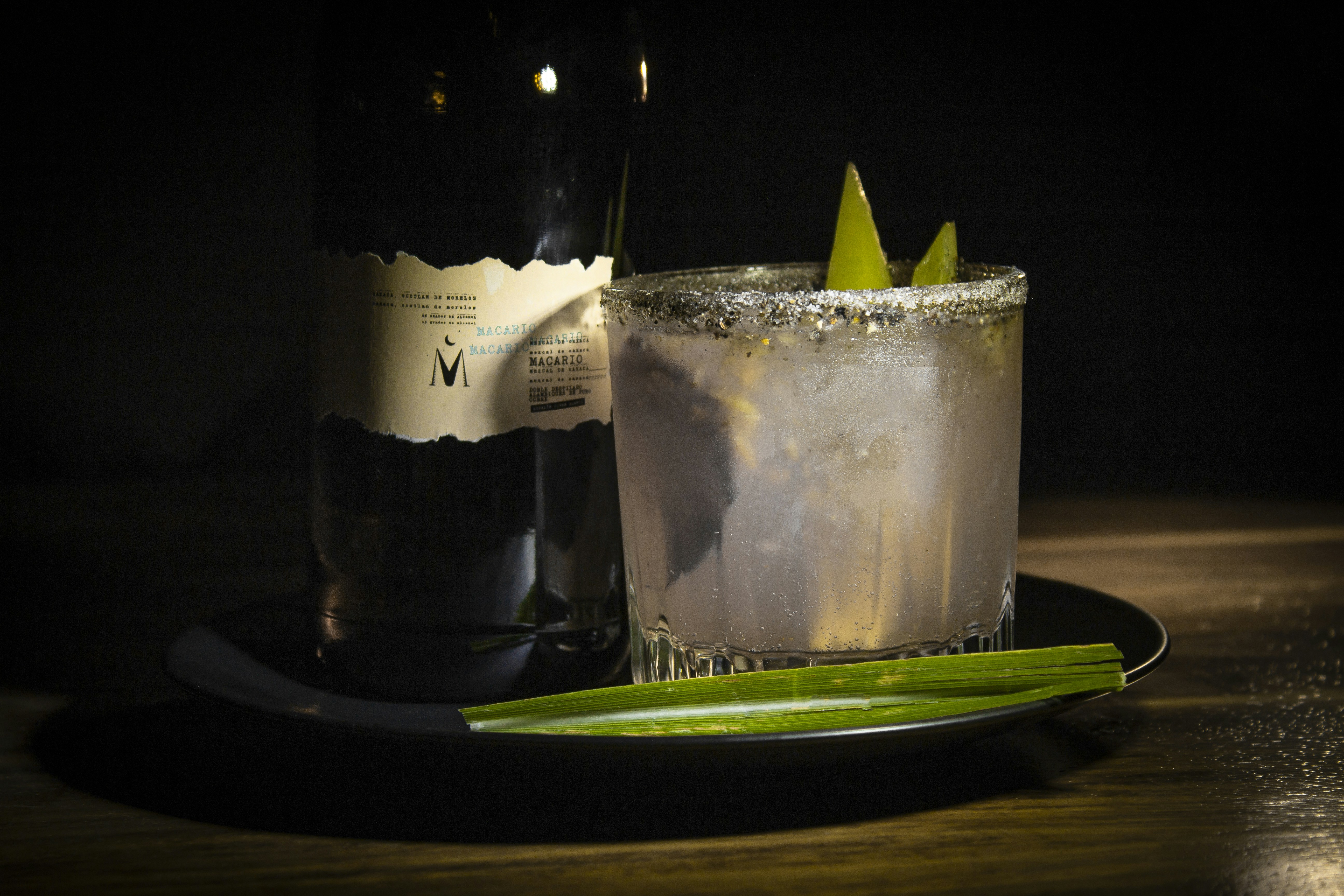 Cocktail glass with lime garnish beside a dark bottle on a wooden table, under dramatic lighting.