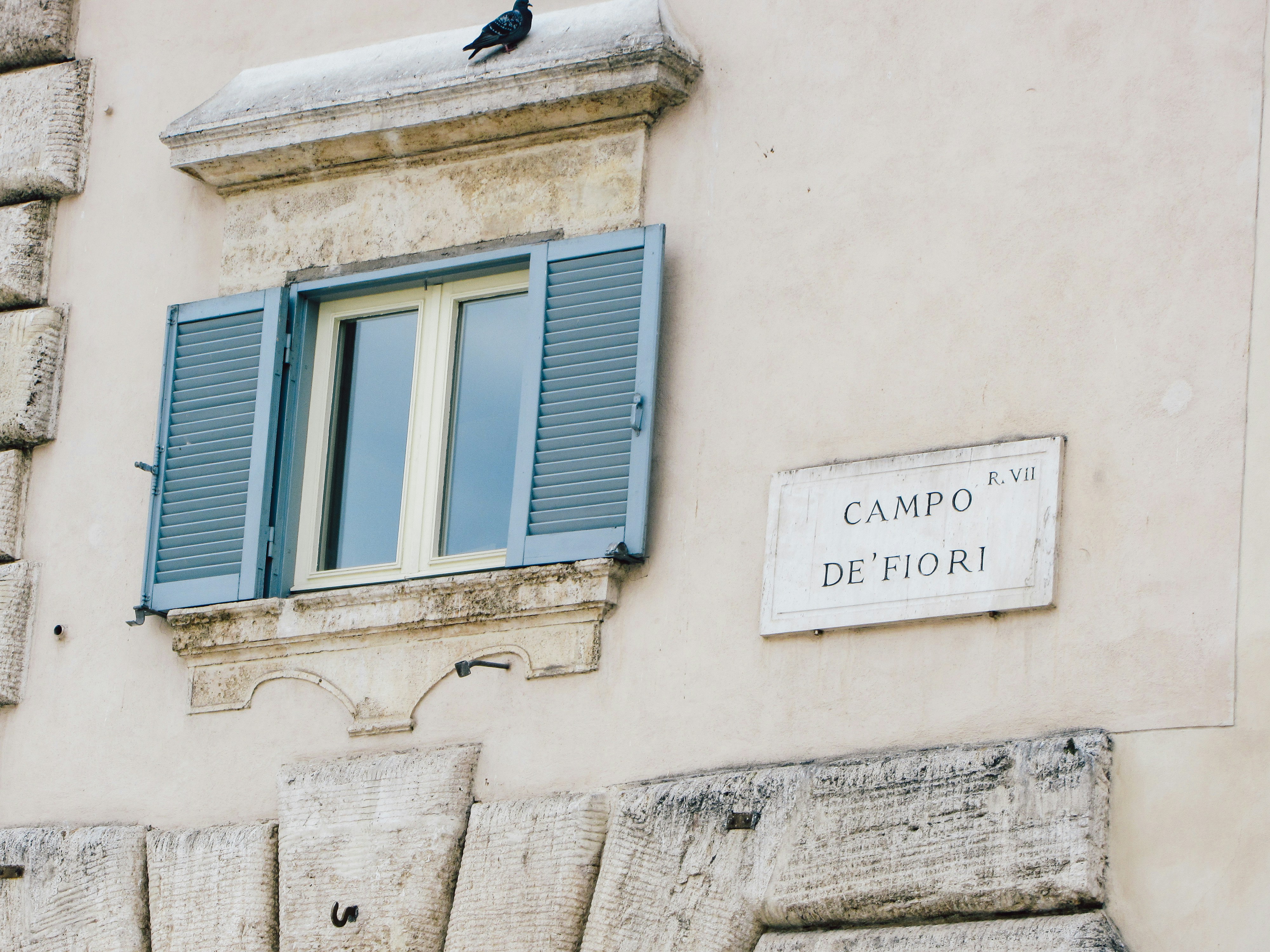 Stone facade featuring a blue-shuttered window and a pigeon perched above, with a Campo de Fiori plaque on the wall.