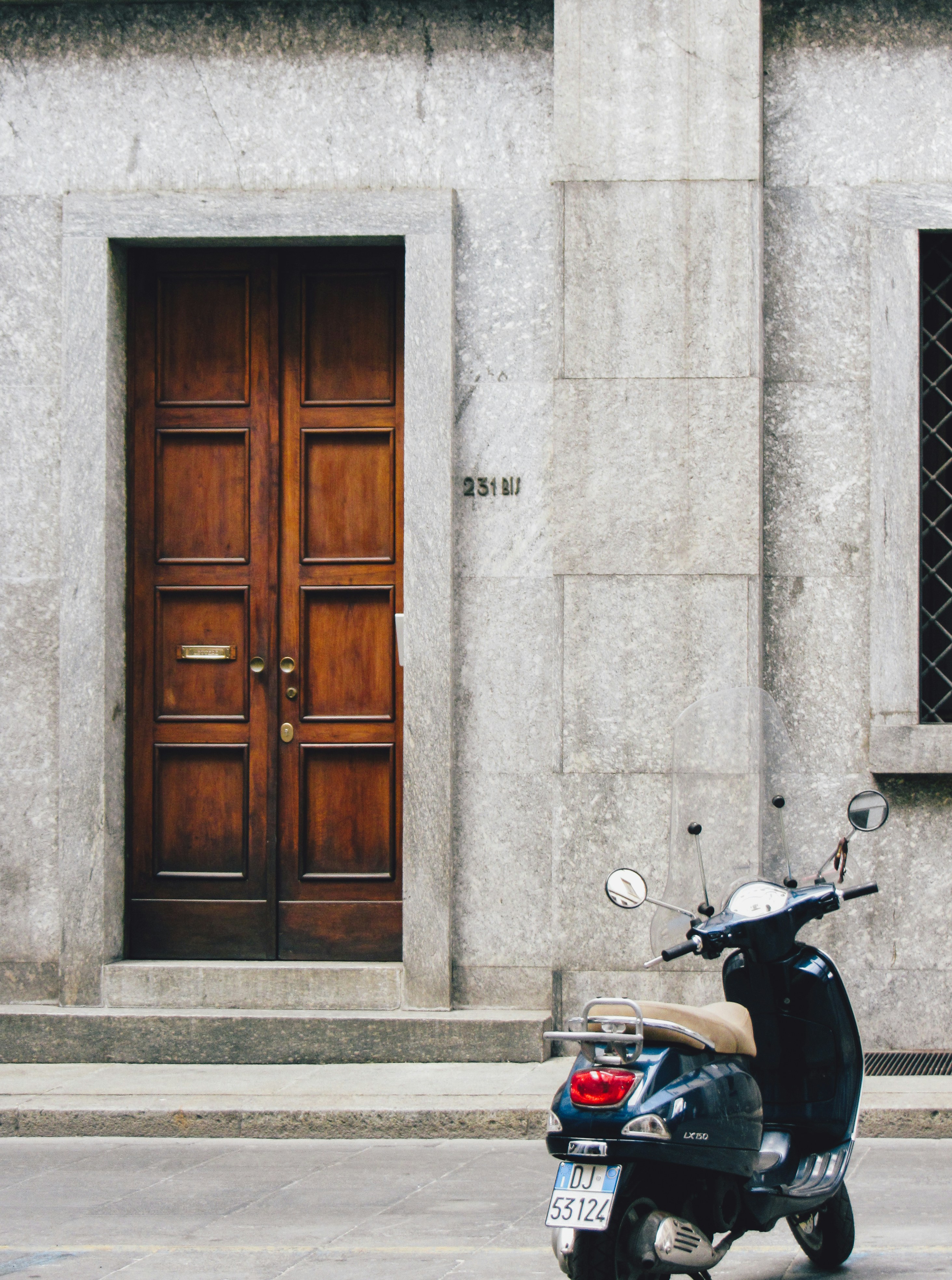 A photograph of a wooden-paneled door set in a gray granite façade, with a scooter parked along the curb. The composition highlights texture and quiet street life.