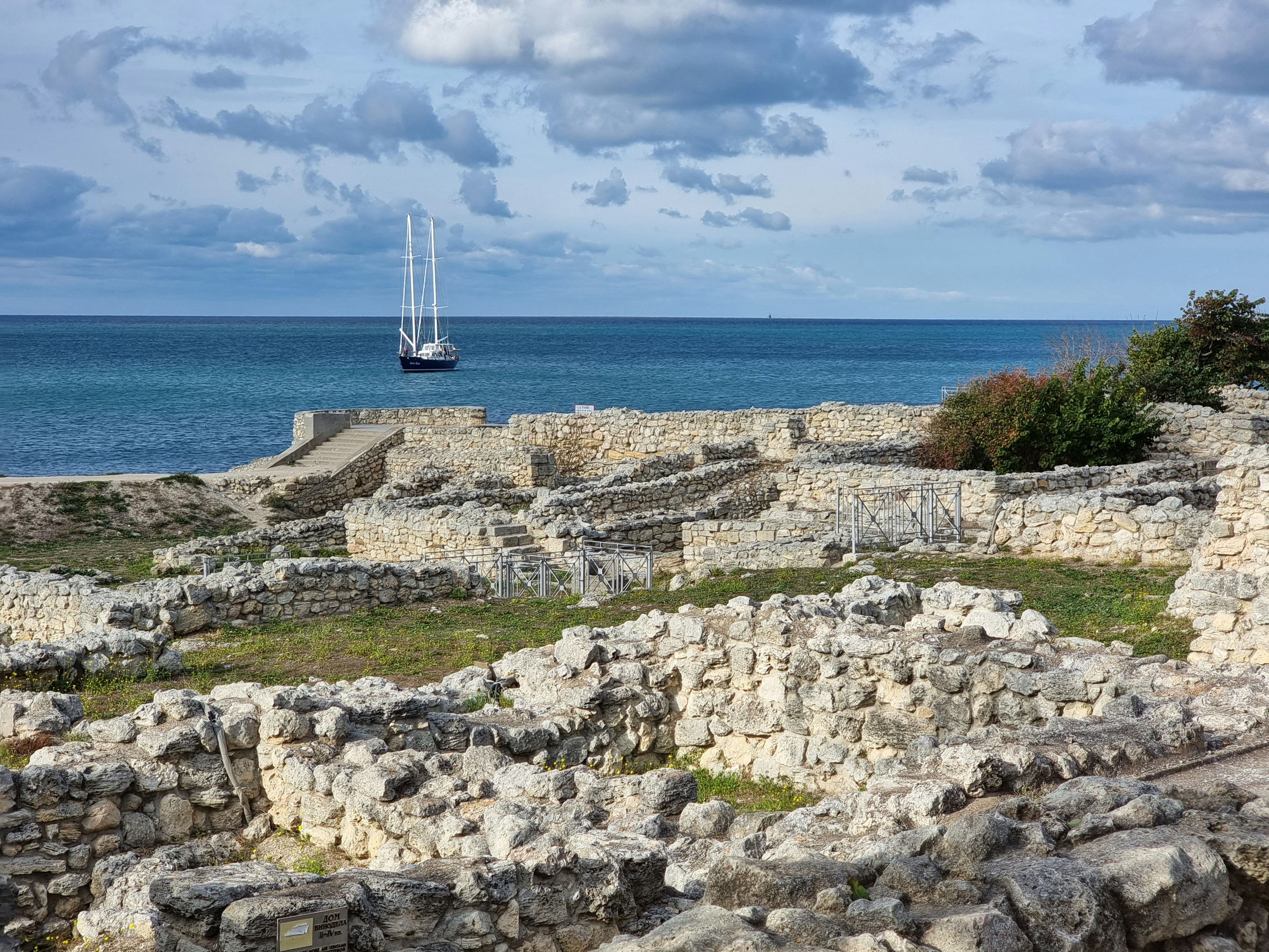 Ancient stone ruins line the foreground along a weathered sea wall. A lone sailboat drifts on the horizon beneath a blue, cloud-dappled sky.