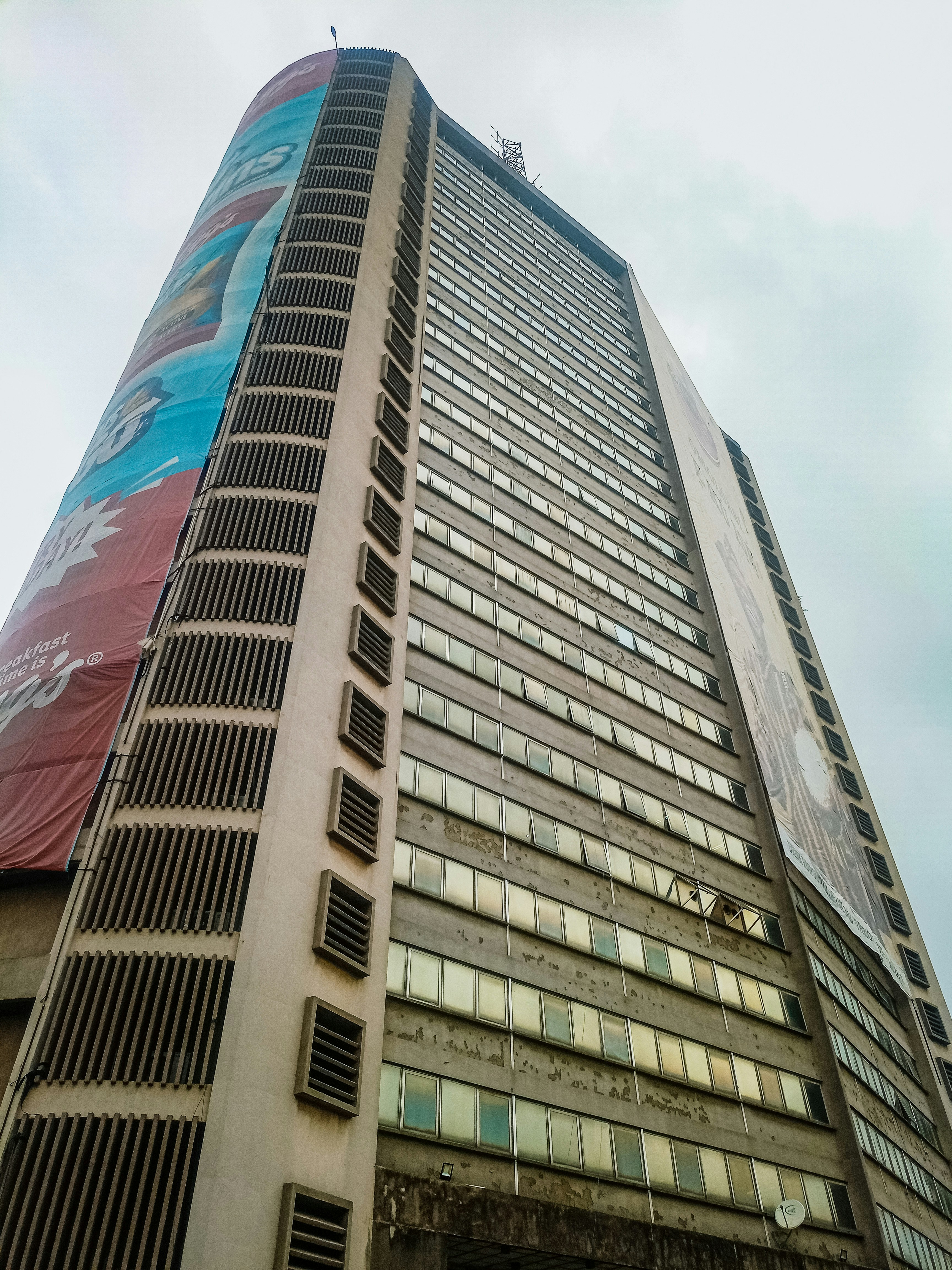 A tall high-rise with rows of windows and colorful banners along the left side, captured from a low-angle against a pale, cloudy sky.
