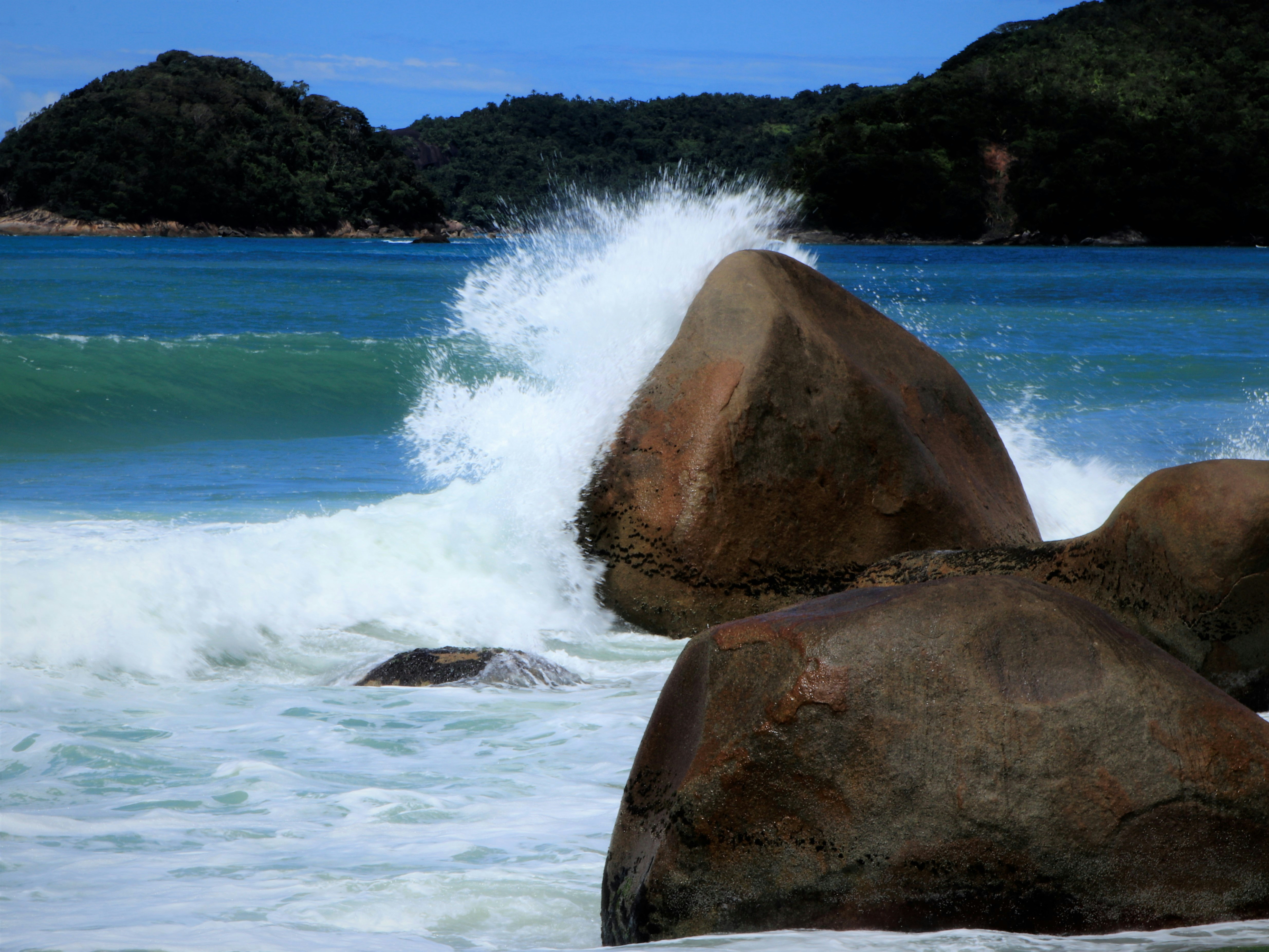 Waves crashing against large rocks along a vibrant coastline under a clear blue sky.