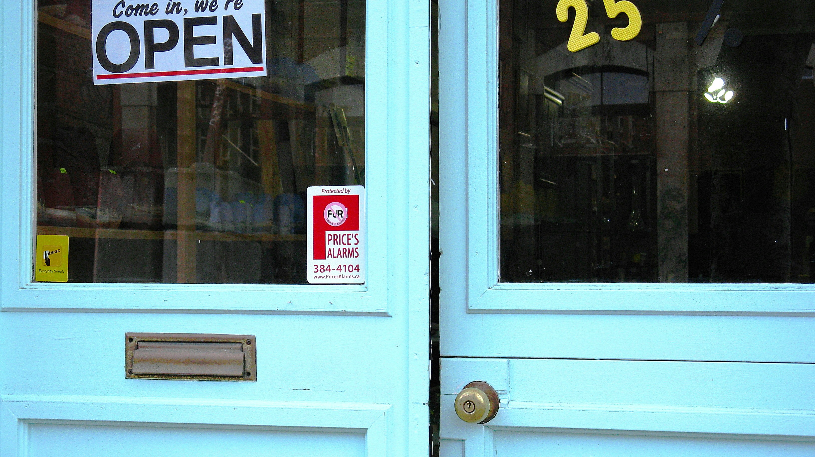 Photograph of bright blue storefront doors framing a glass entrance, with an OPEN sign and a security alarm sticker visible in the left pane. A brass doorknob and mail slot anchor the lower half.
