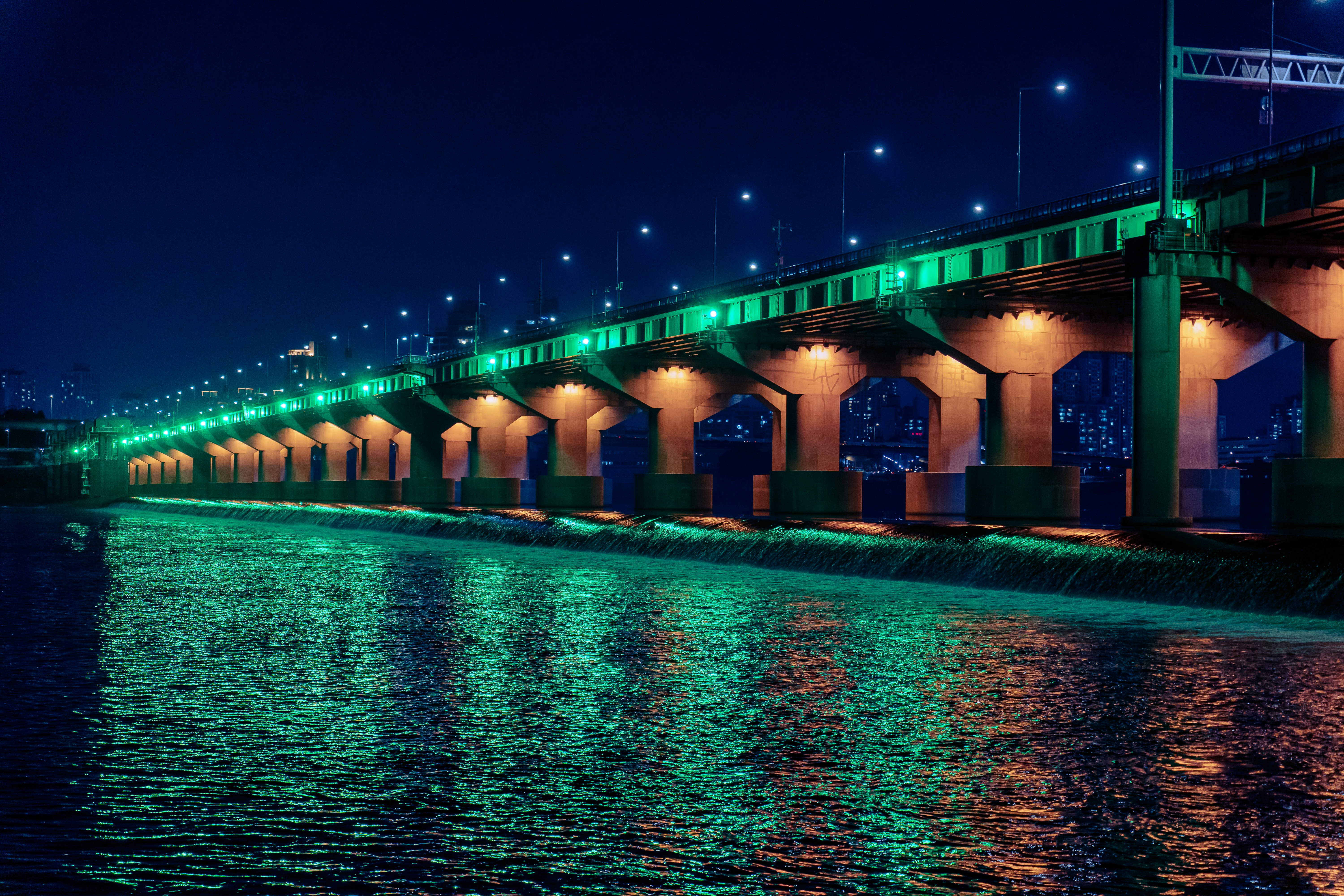 Illuminated bridge at night, casting vibrant reflections on the water below. The scene captures the interplay of light and shadow in an urban landscape.