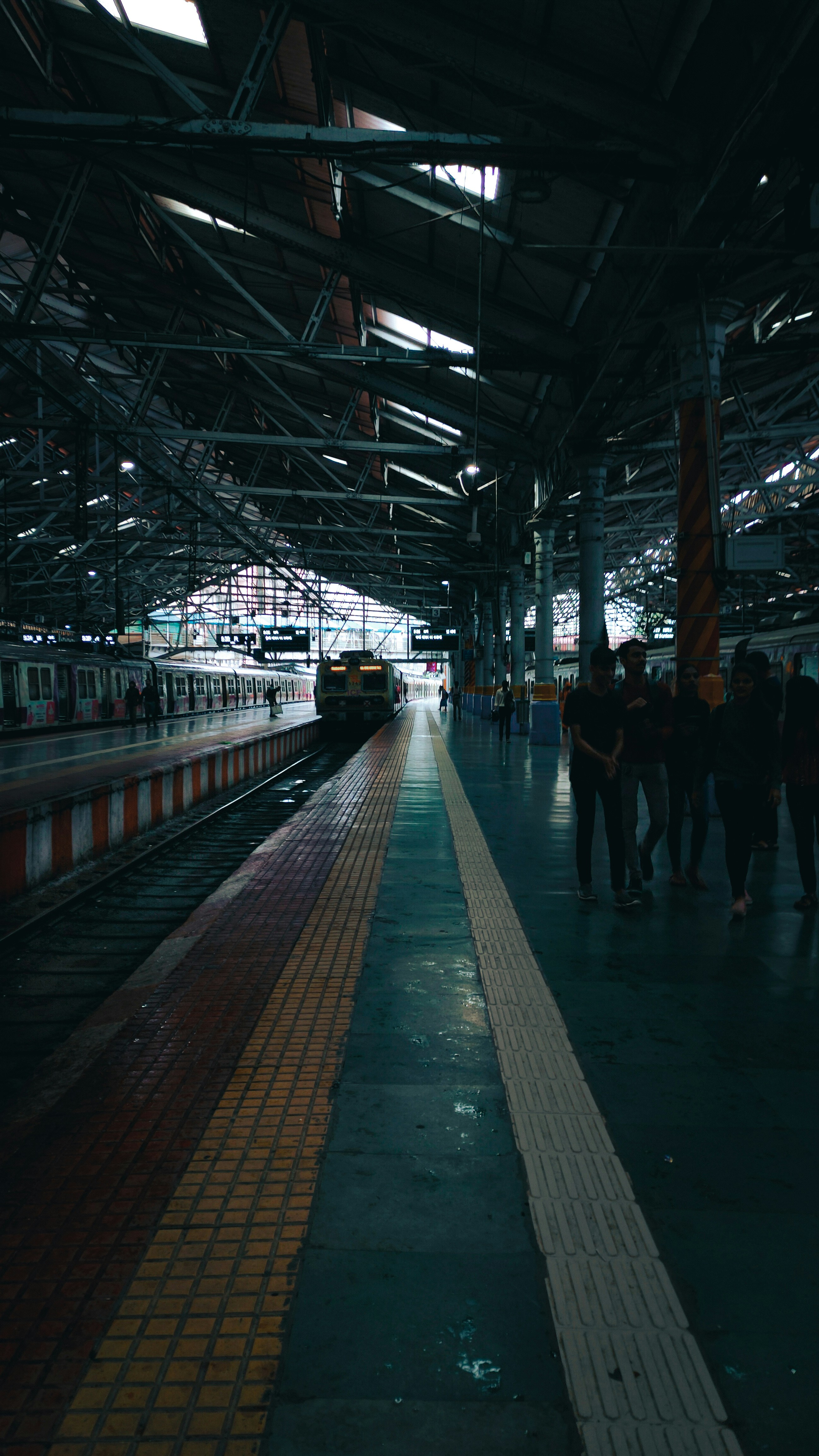Dimly lit train platform photograph with a distant train approaching along the tracks. Passengers stand along the right, while a network of steel beams and skylights looms overhead.