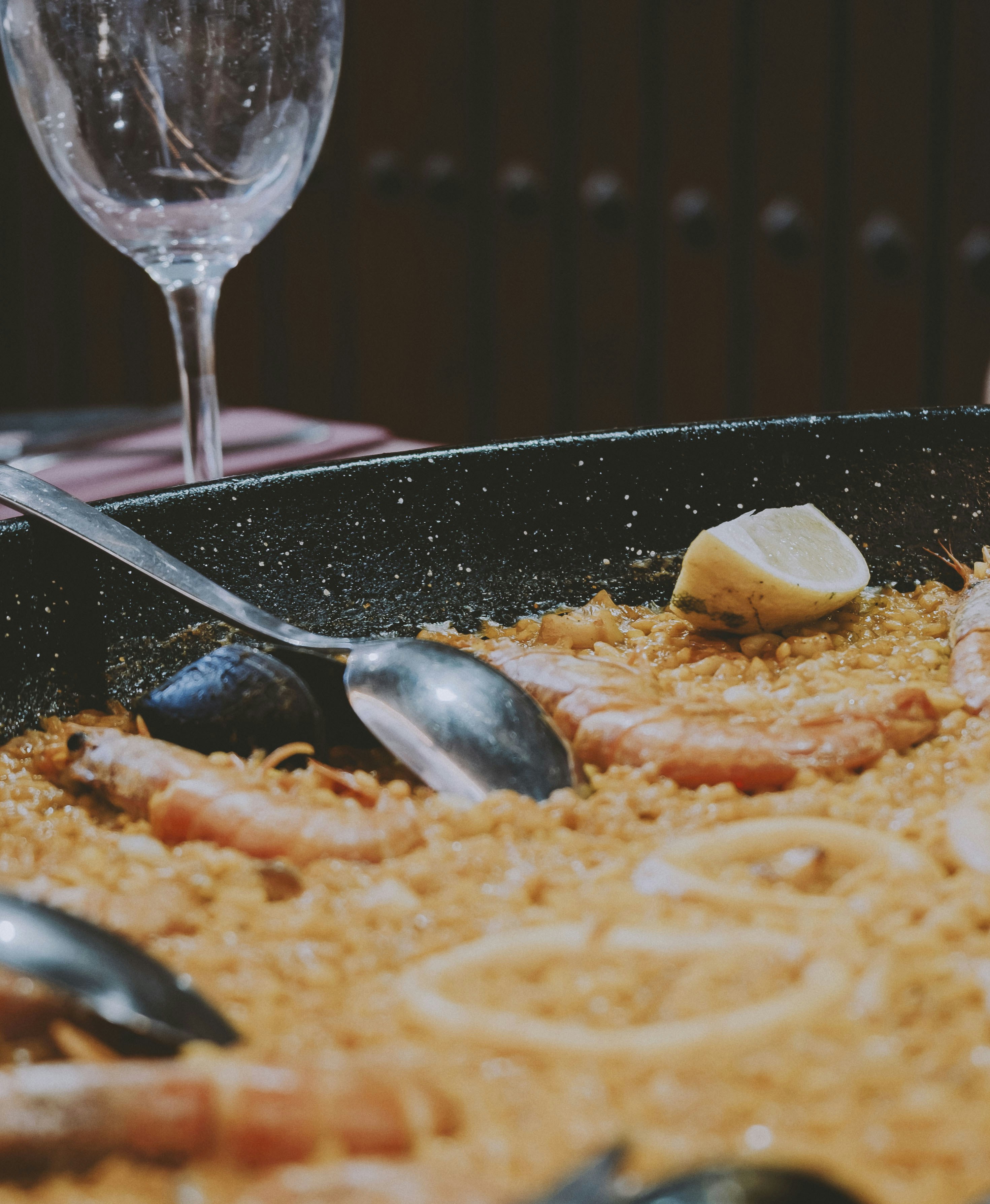 A vibrant paella dish featuring shrimp, mussels, and lemon slices, served in a traditional pan. A glass of wine adds to the dining experience.