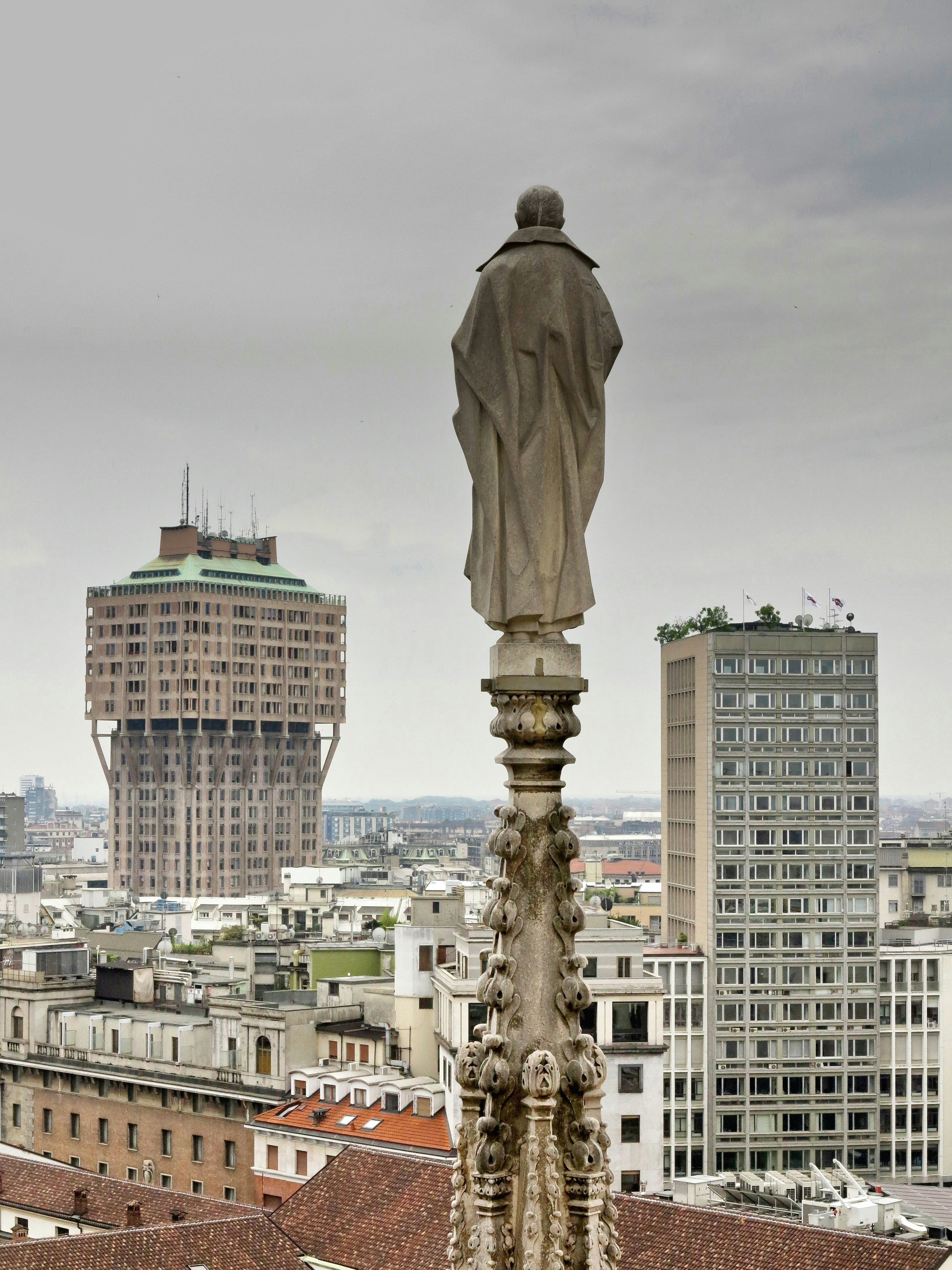 Stone statue atop a tall, ornate spire overlooks a dense city skyline. The scene juxtaposes historic sculpture with modern architecture.