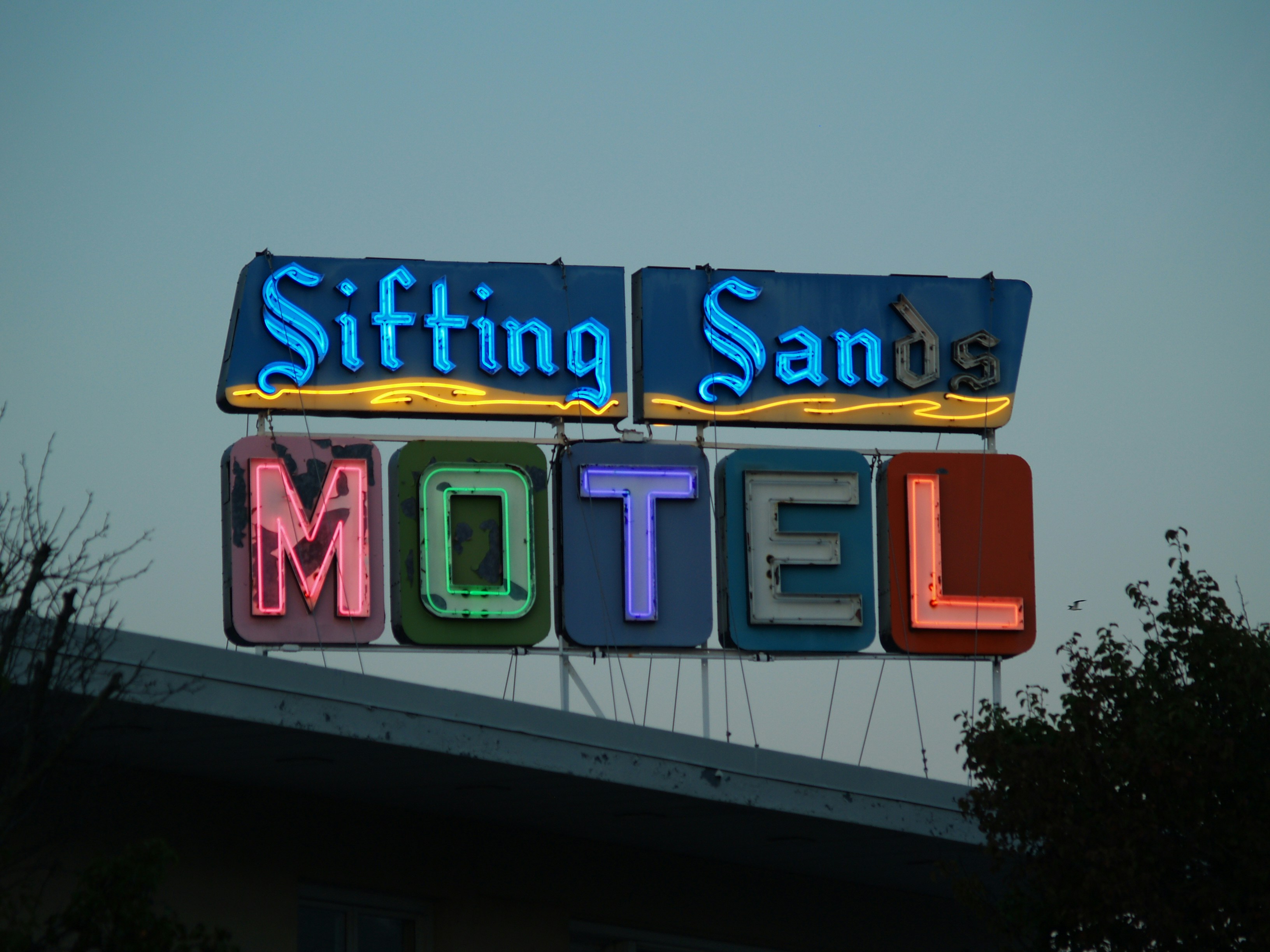 Vibrant neon sign for Sifting Sands Motel, illuminated against a twilight sky, showcasing retro charm and inviting travelers. 