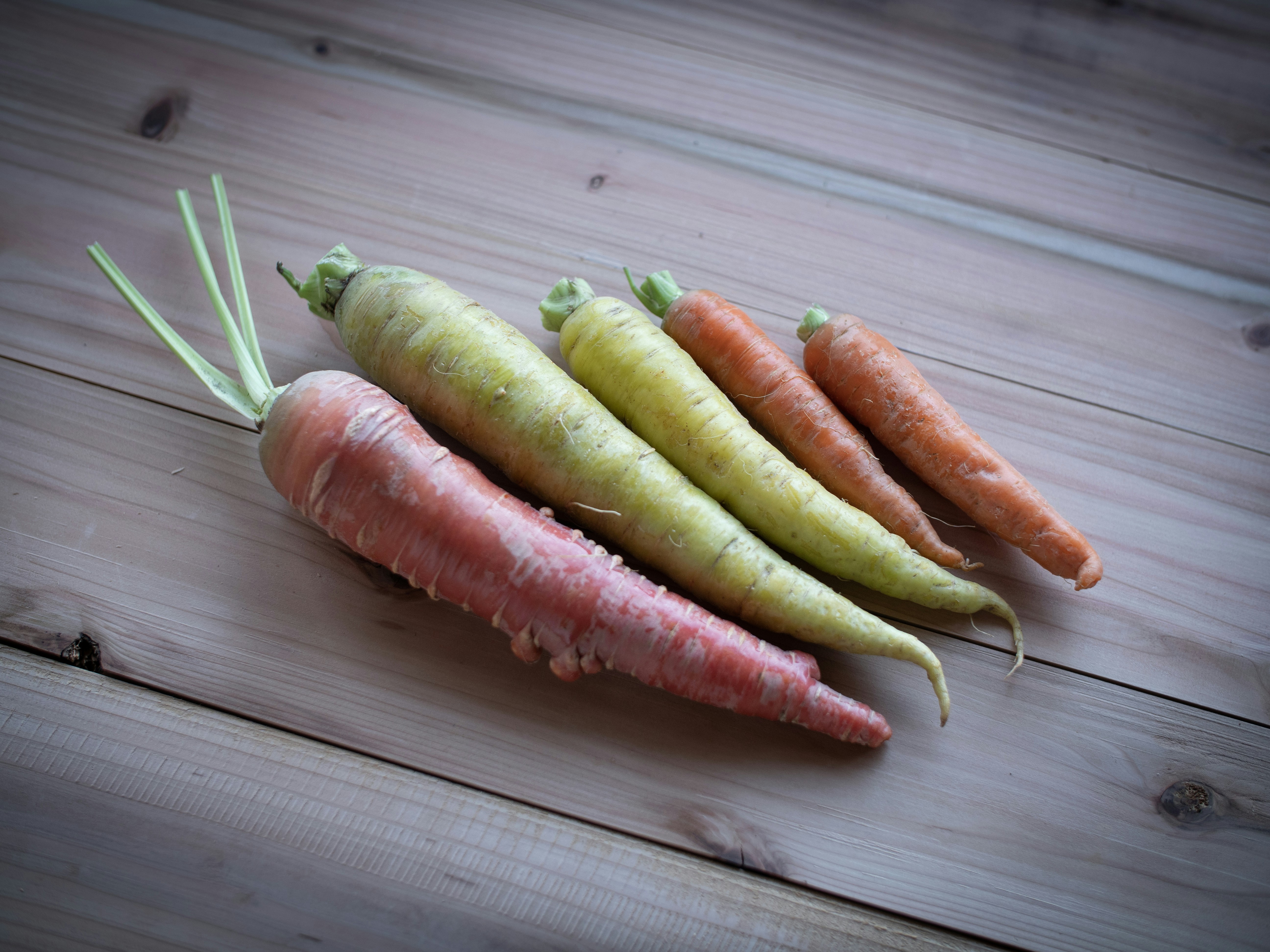 Row of fresh, multicolored carrots resting on a light wooden surface, showcasing varied hues from pink to yellow. This is a still-life photograph of produce on a rustic tabletop.