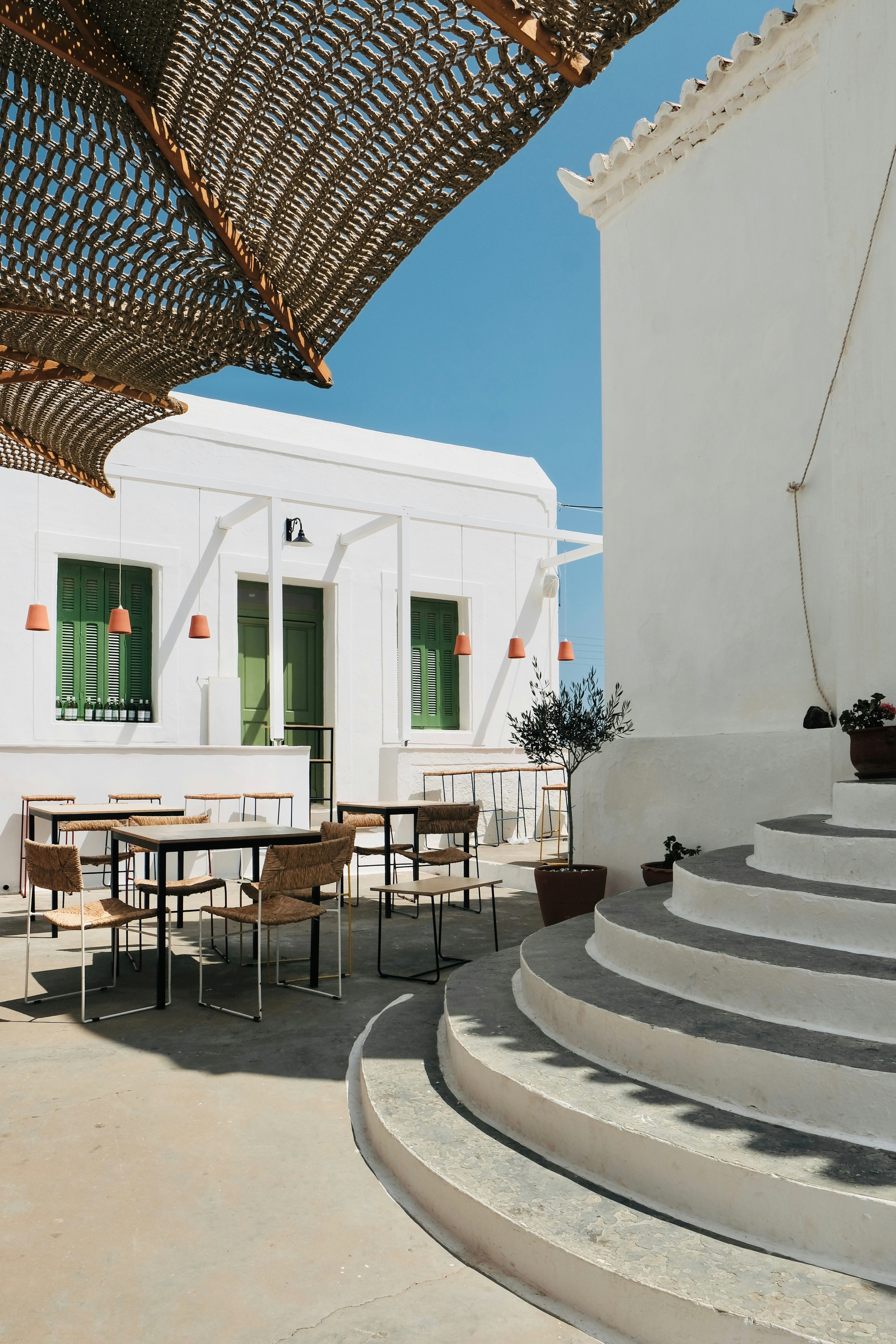 Charming outdoor café space featuring wooden tables and steps leading to a white building under a woven shade structure. Potted plants add a touch of greenery.