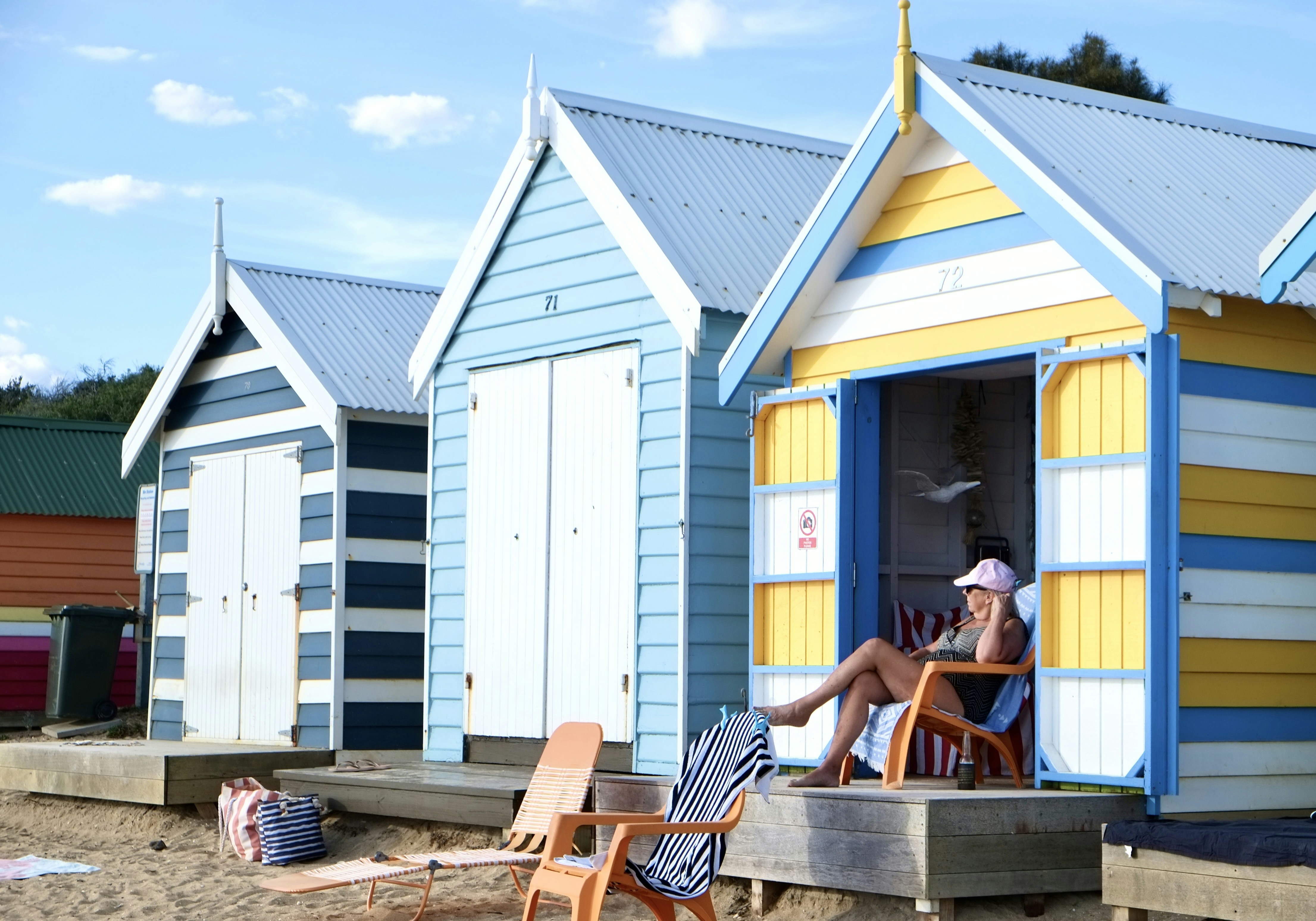 A woman relaxes on a deck chair outside a vibrant beach hut, enjoying the sunlit coastal atmosphere. The scene features colorful beach cabins with an inviting ambiance.