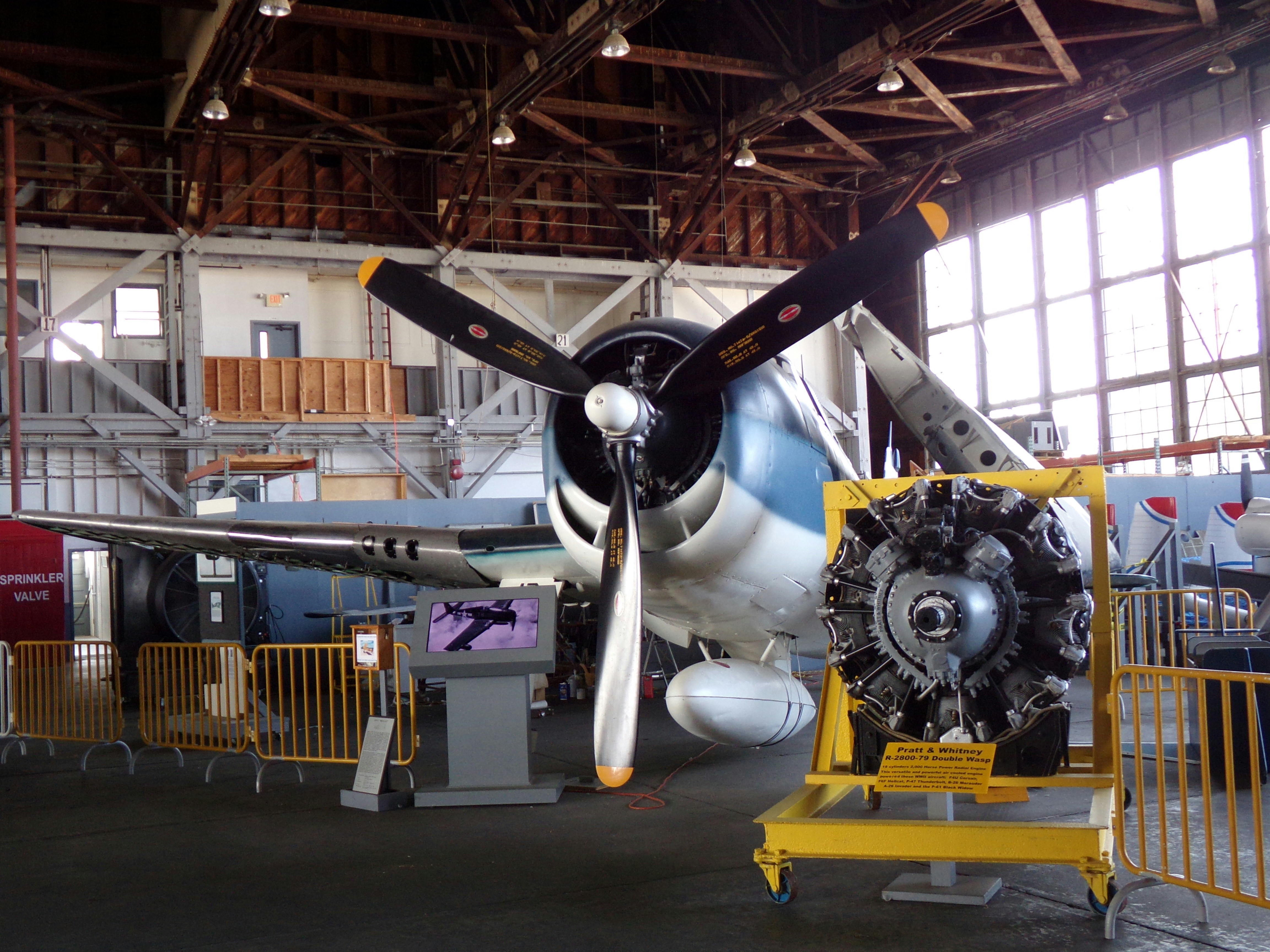 Photograph of a WWII-era propeller aircraft on display, with a disassembled engine on a yellow stand nearby.