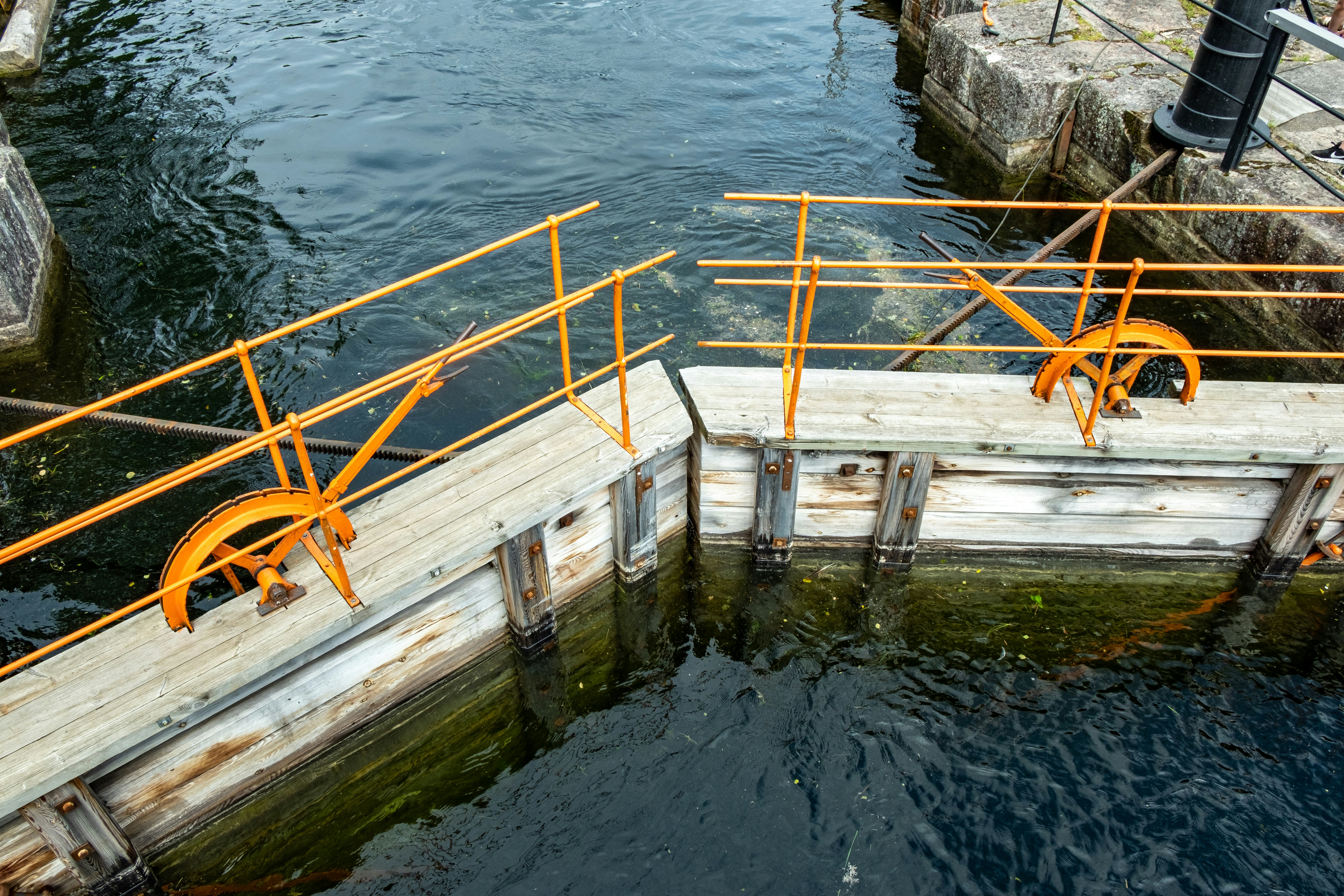 Orange mechanical components on a wooden structure control water flow in a canal, surrounded by dark water and stone edges.