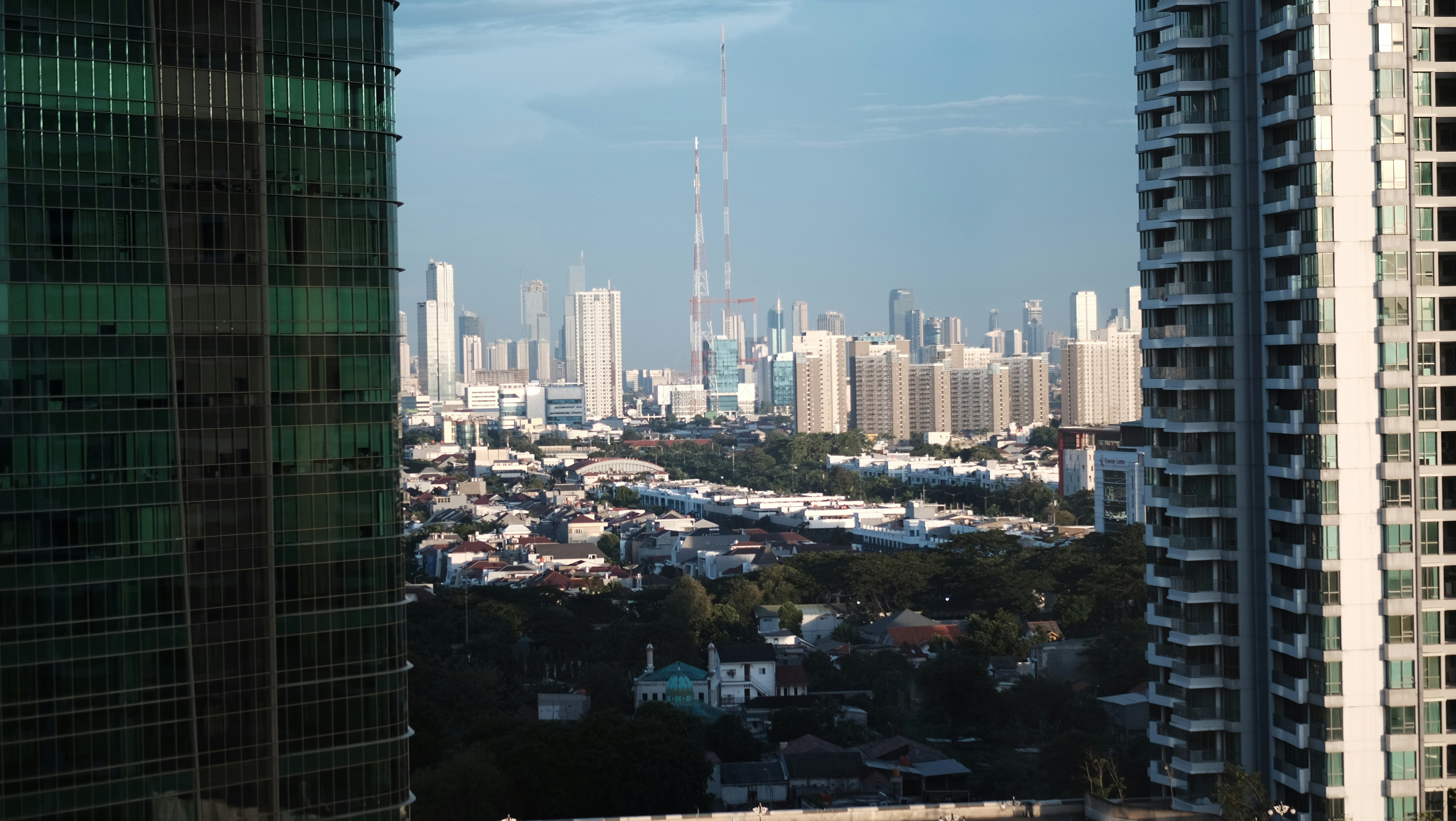 A panoramic view of a bustling city skyline, framed by modern high-rises and lush greenery below. The scene captures the juxtaposition of urban architecture and nature.