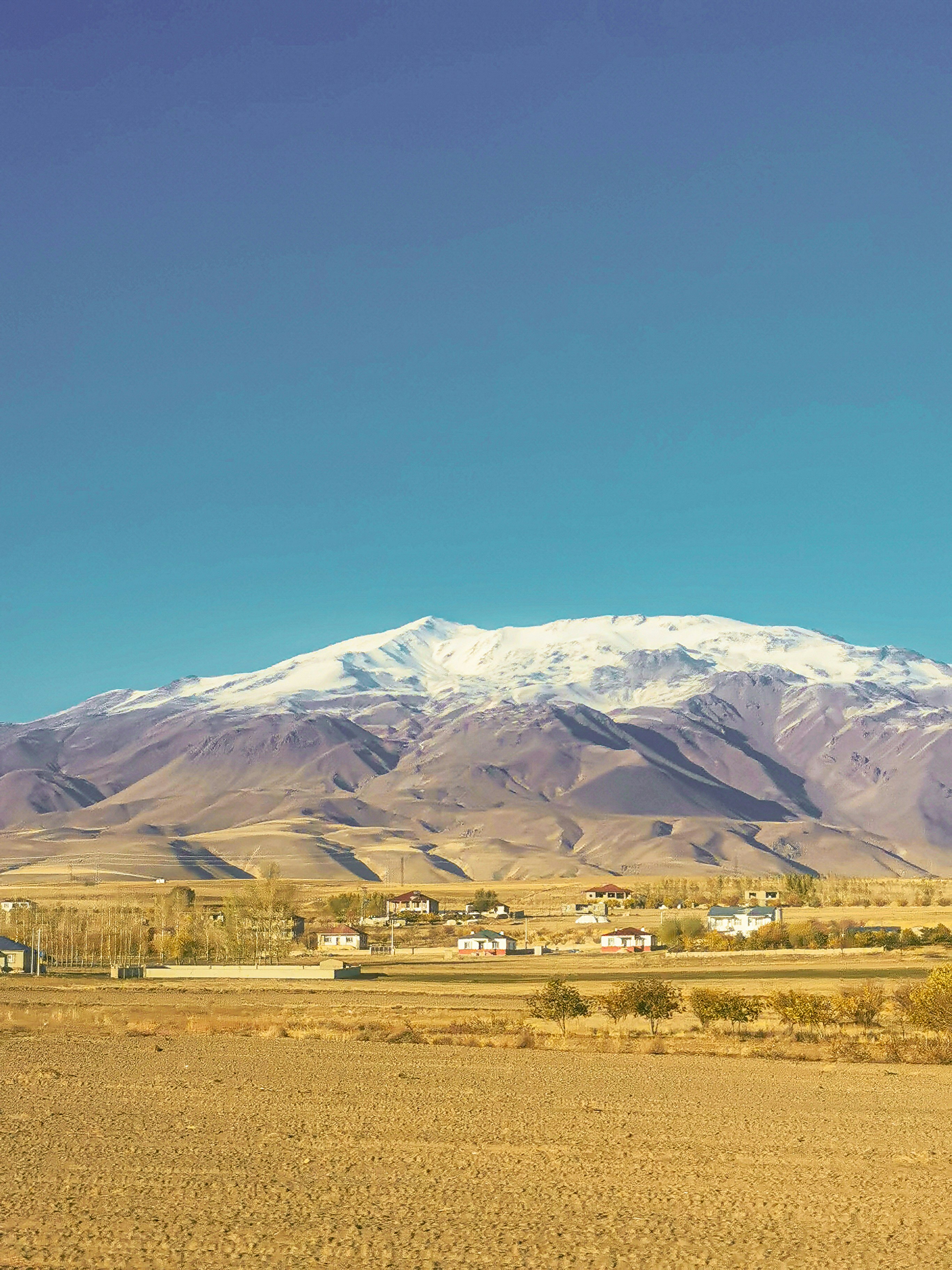 Snow-capped mountain rises above a sunlit valley with scattered buildings and fields.