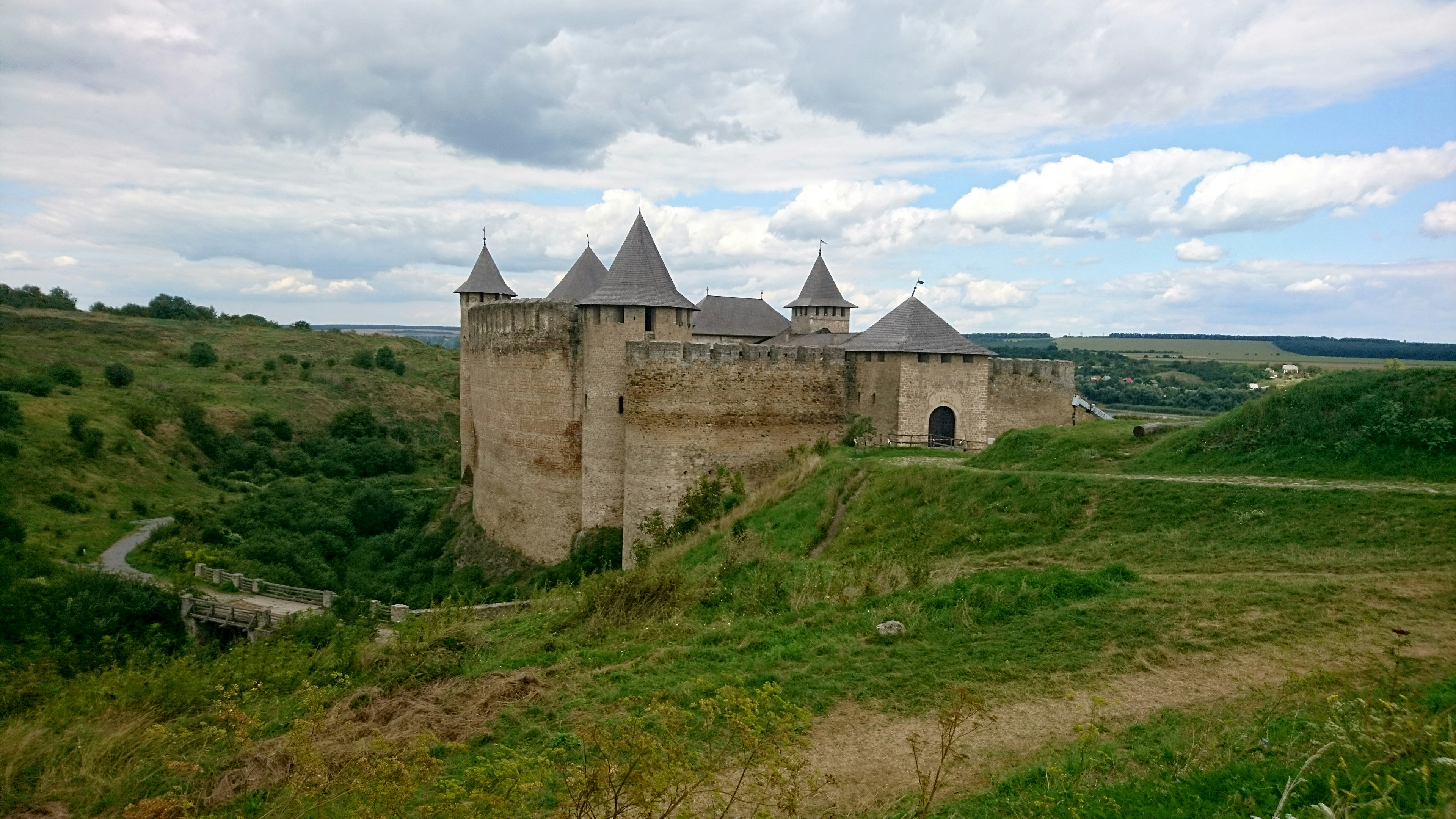 Historic fortress perched on a hill, surrounded by lush greenery and a winding path. The structure features distinctive towers and a rugged stone facade.