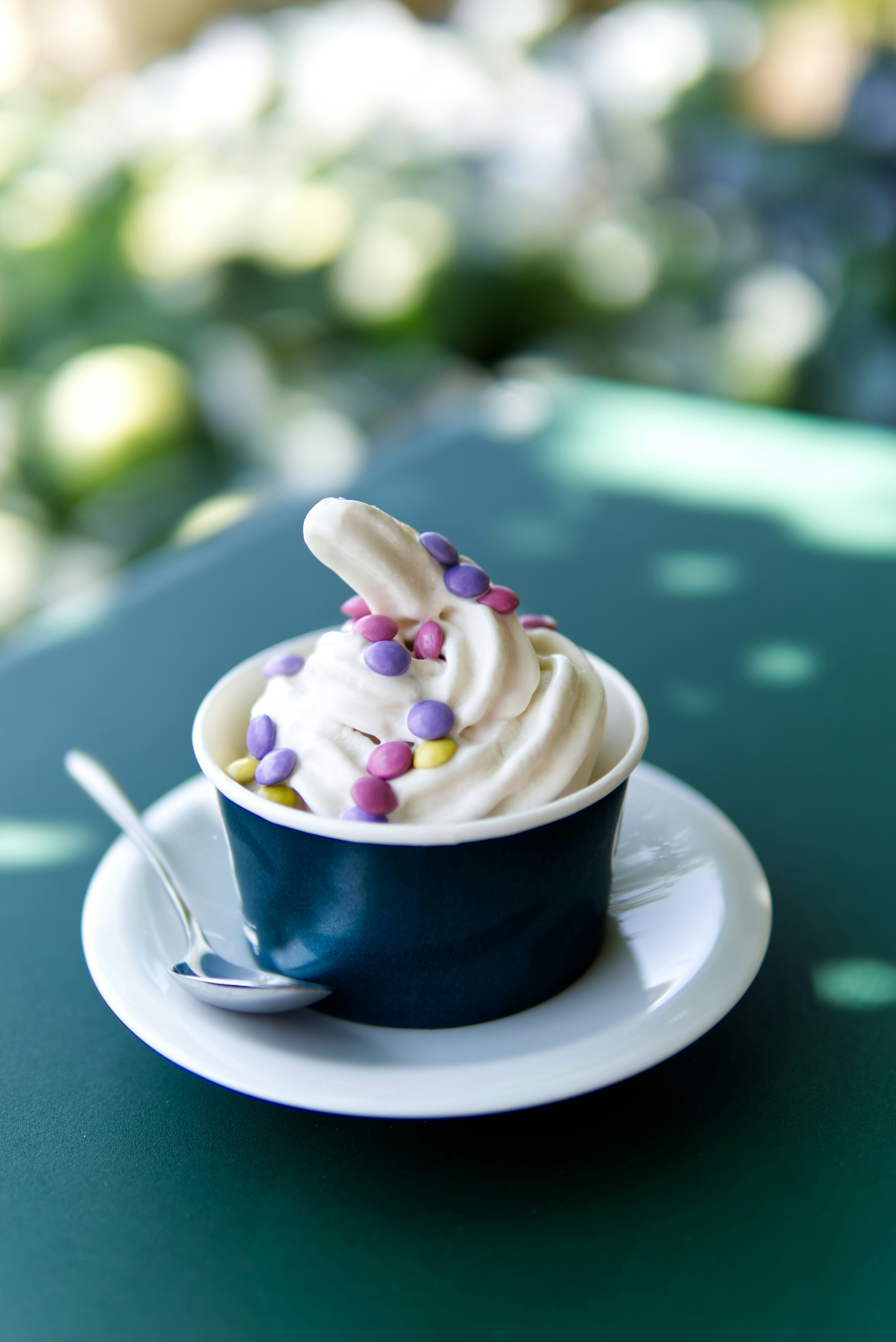 Soft-serve ice cream adorned with colorful candy pieces, served in a dark cup on a white plate.