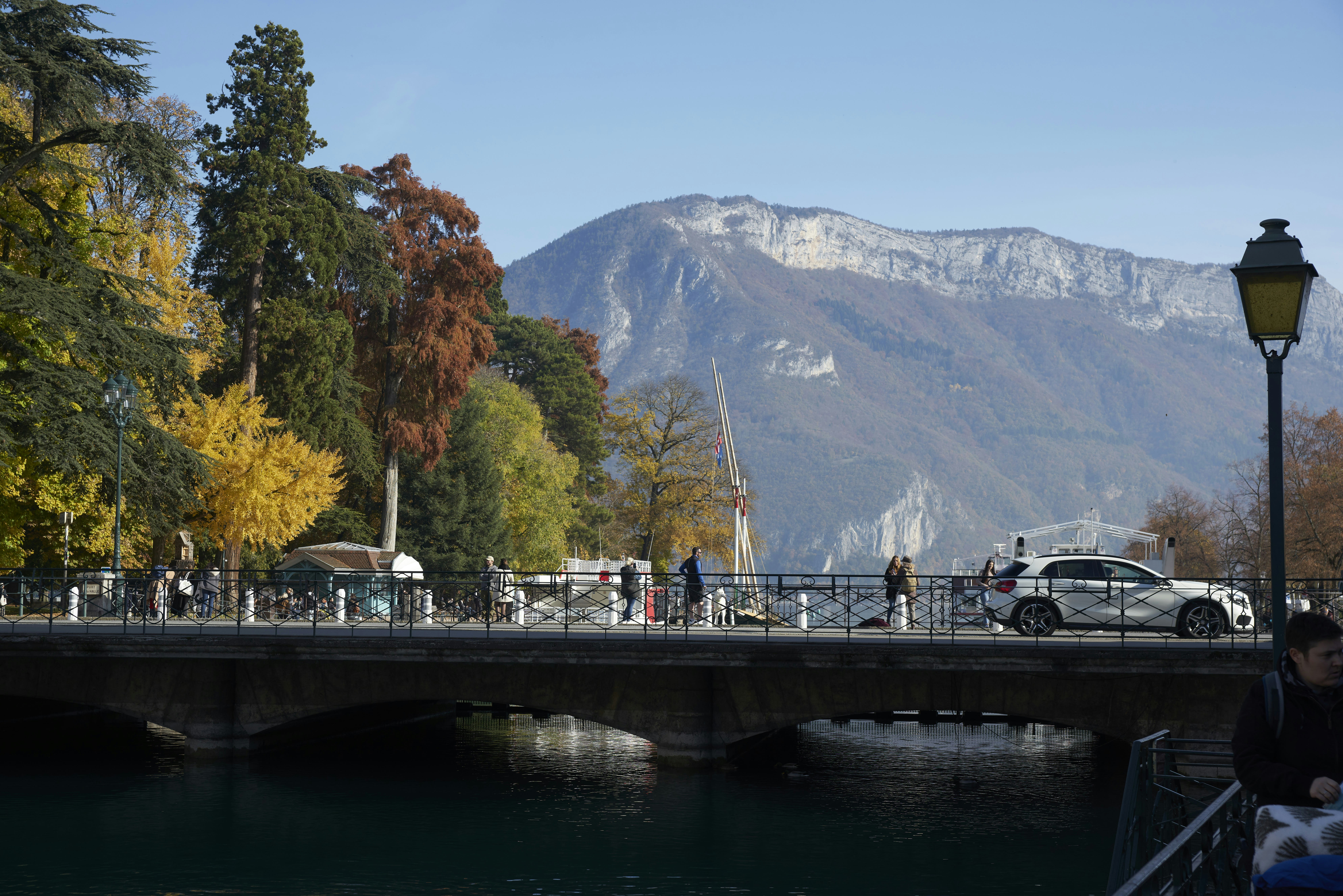 Pedestrians and a car cross a bridge over a calm river, framed by autumn trees with distant snow-dusted Alps.