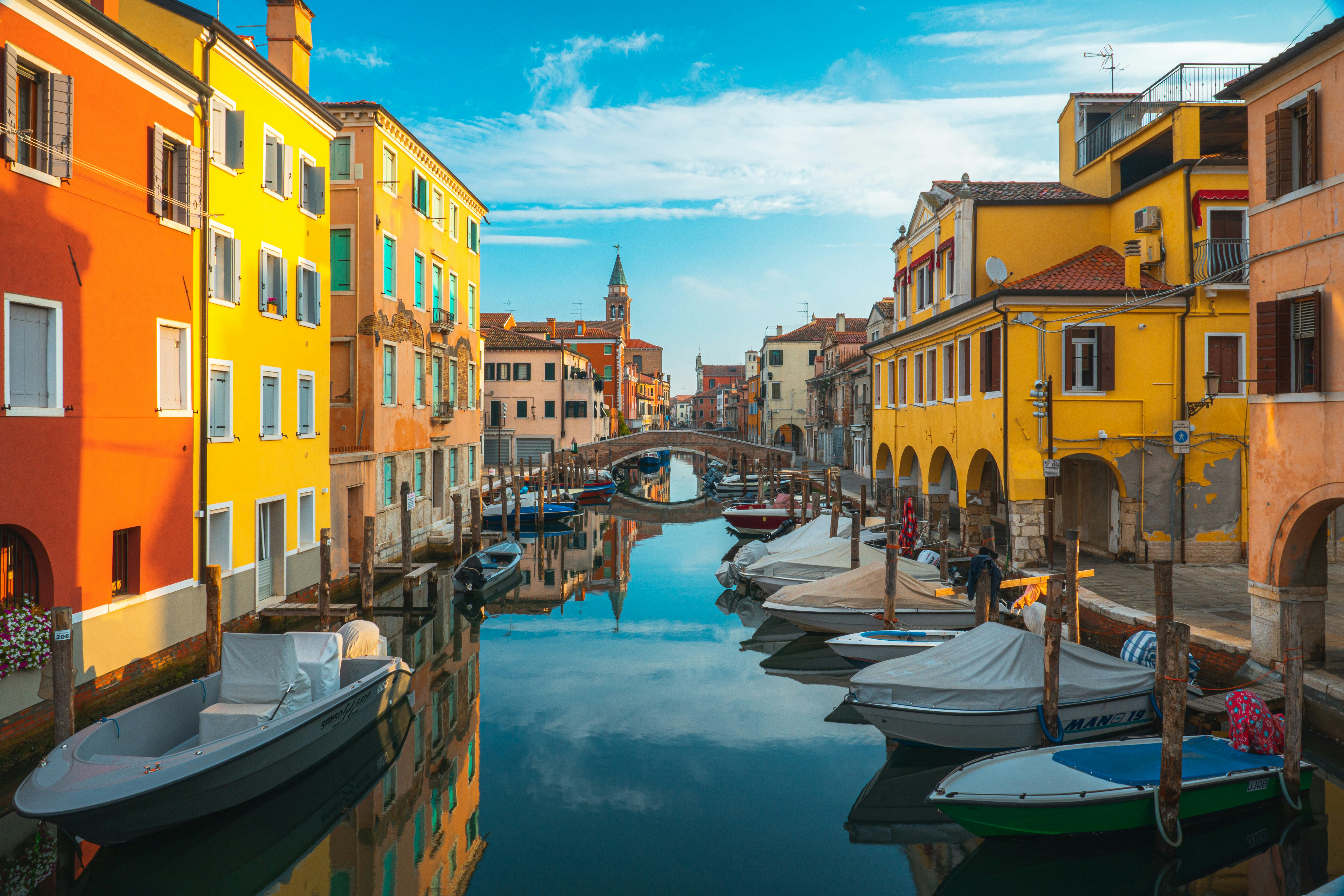 Colorful buildings line a tranquil canal with moored boats under a bright blue sky.