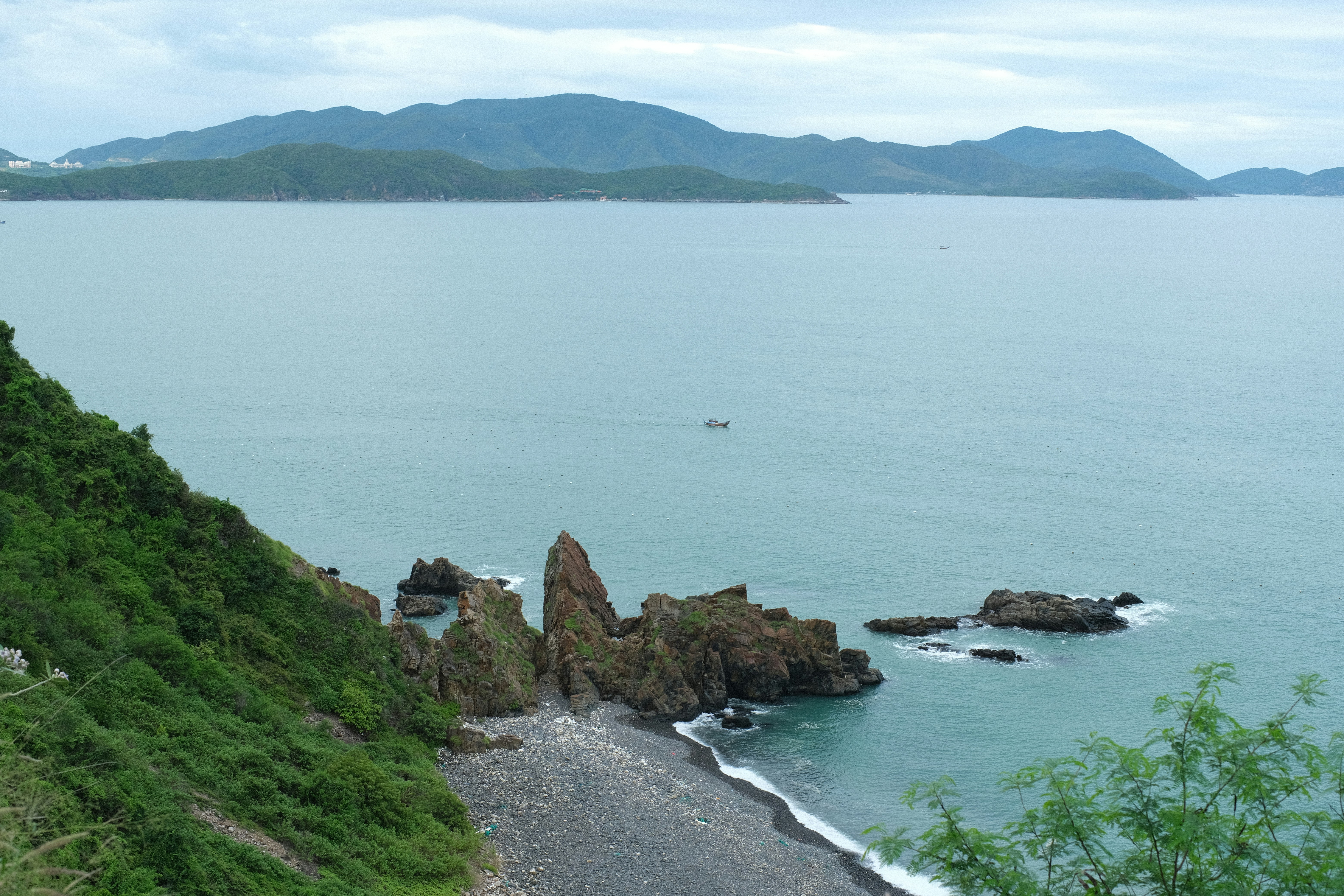 Rocky shoreline meeting calm sea under an overcast sky, with distant mountains on the horizon.