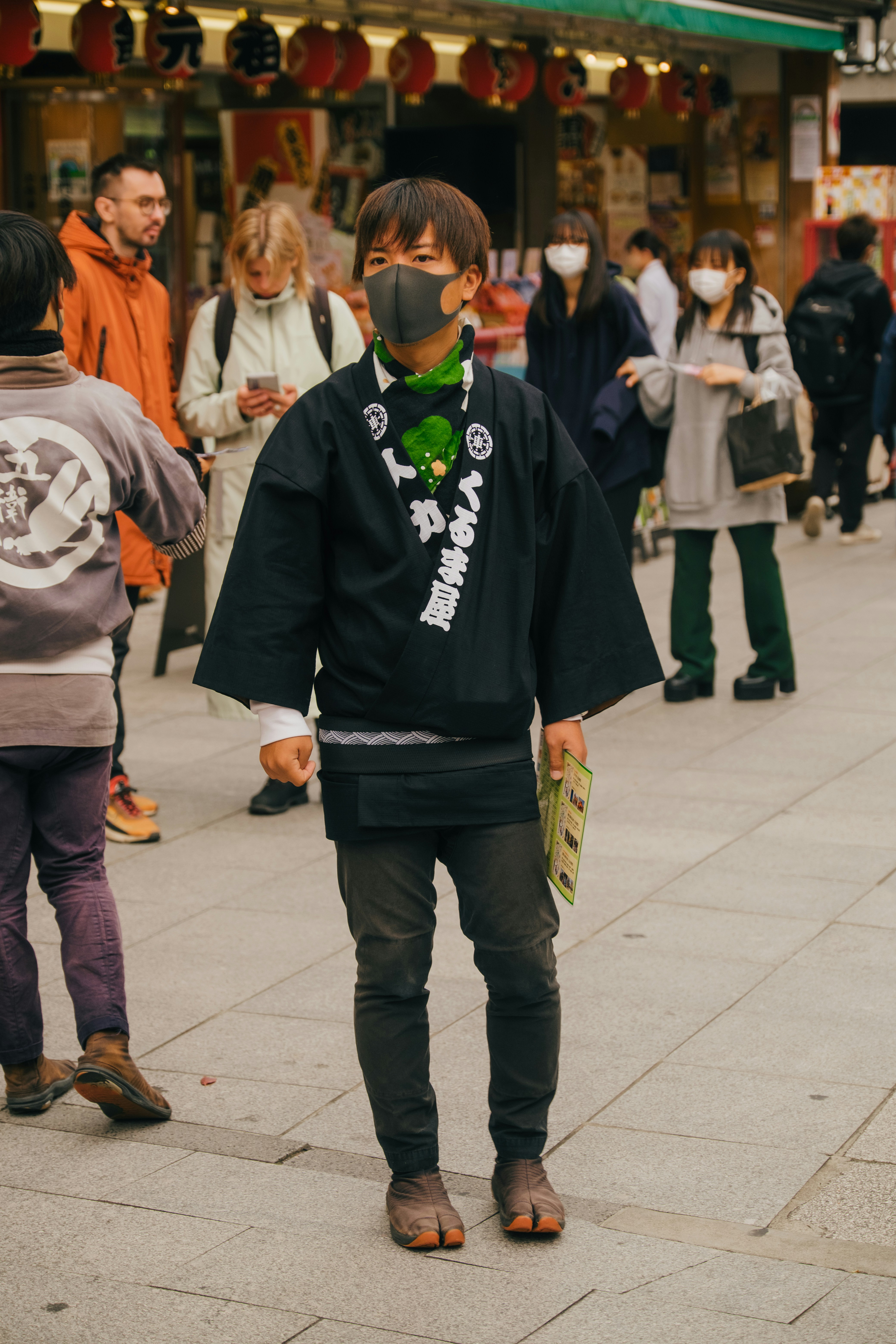 Lantern-lit street: a masked performer in a black happi coat stands center amid a bustling crowd