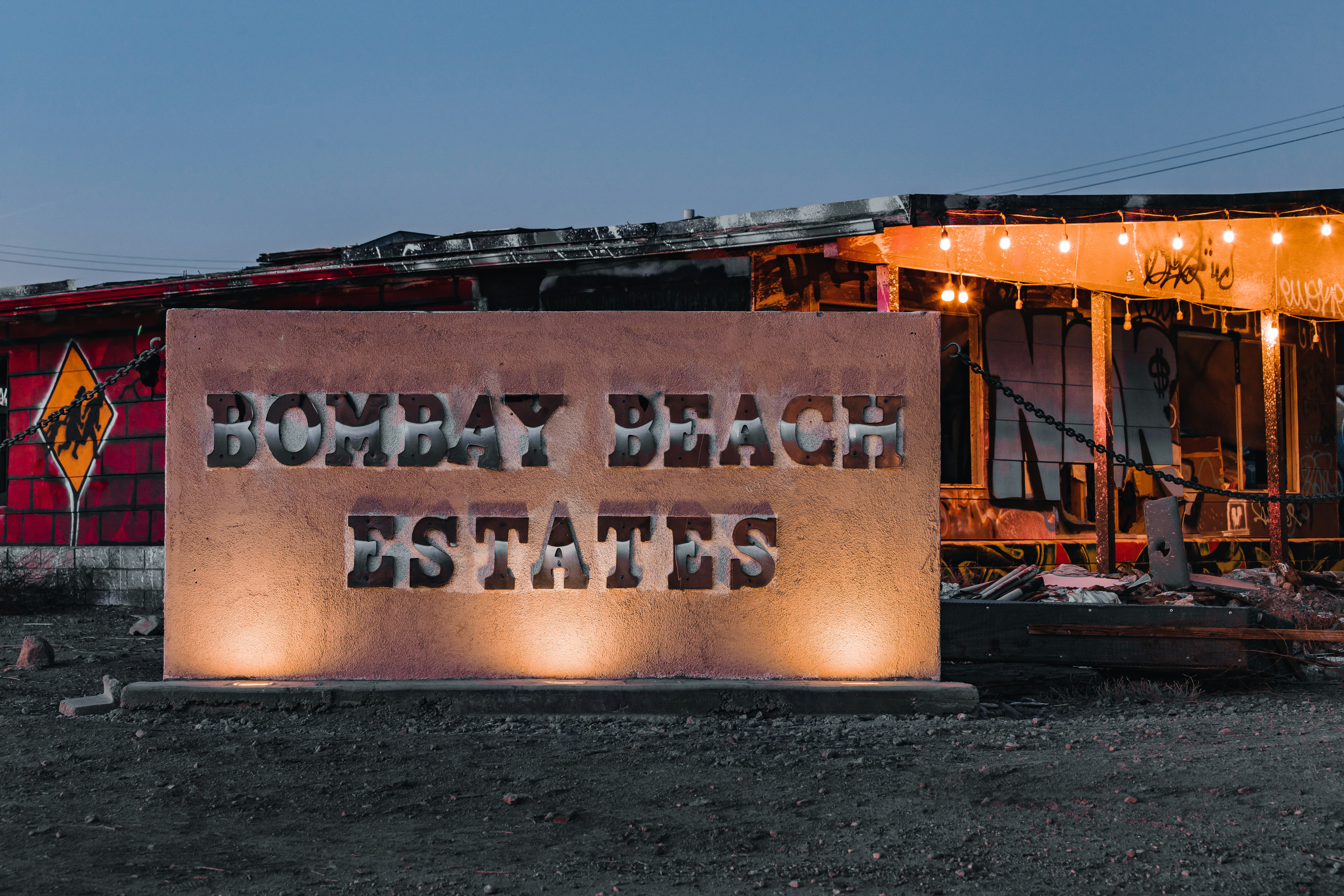 Illuminated Bombay Beach Estates sign at dusk, with a rugged, rustic building facade in the background and string lights along the top edge.