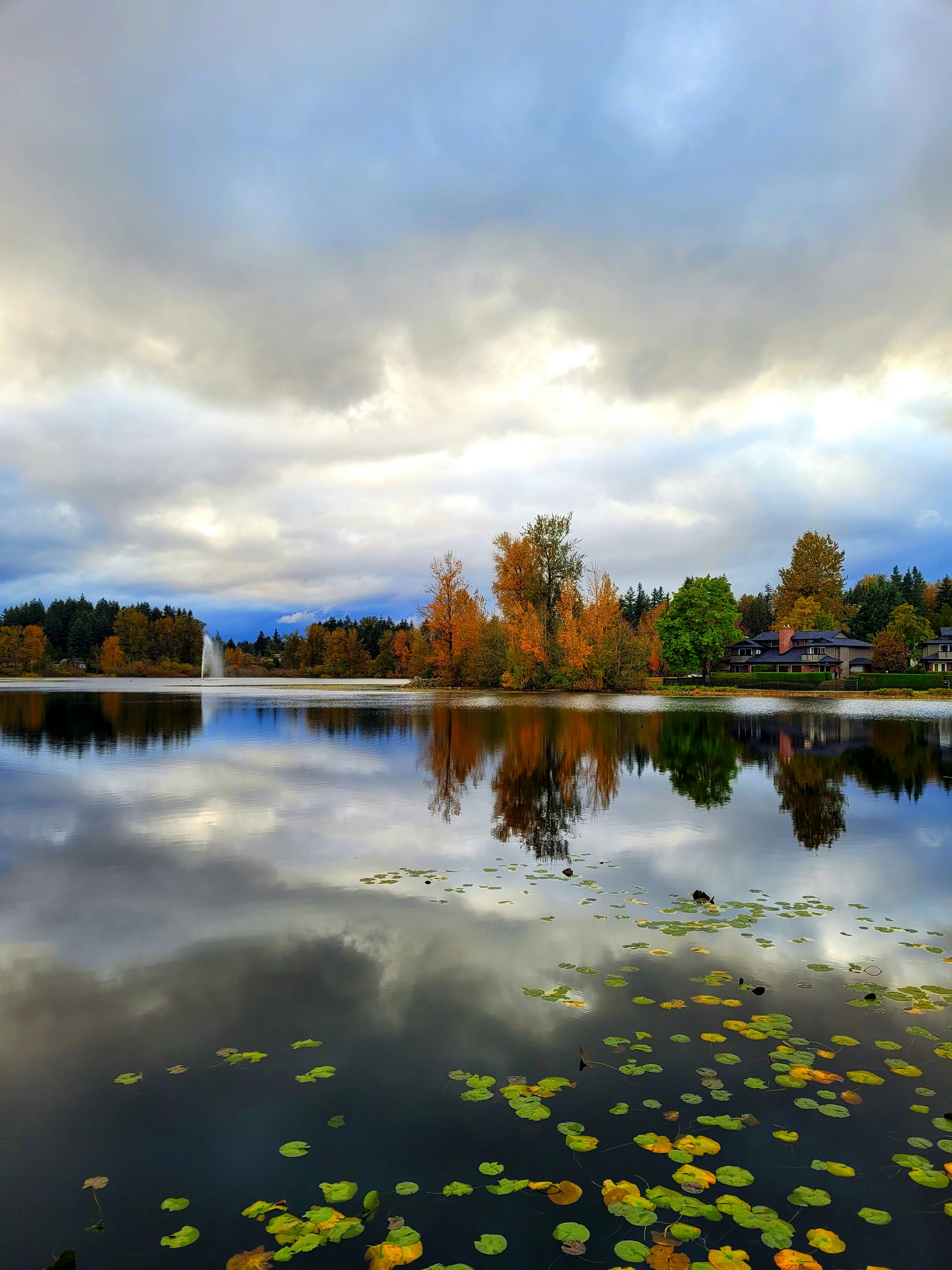 A tranquil lake scene reflecting vibrant autumn foliage under a moody sky, with a fountain adding a dynamic element to the landscape.