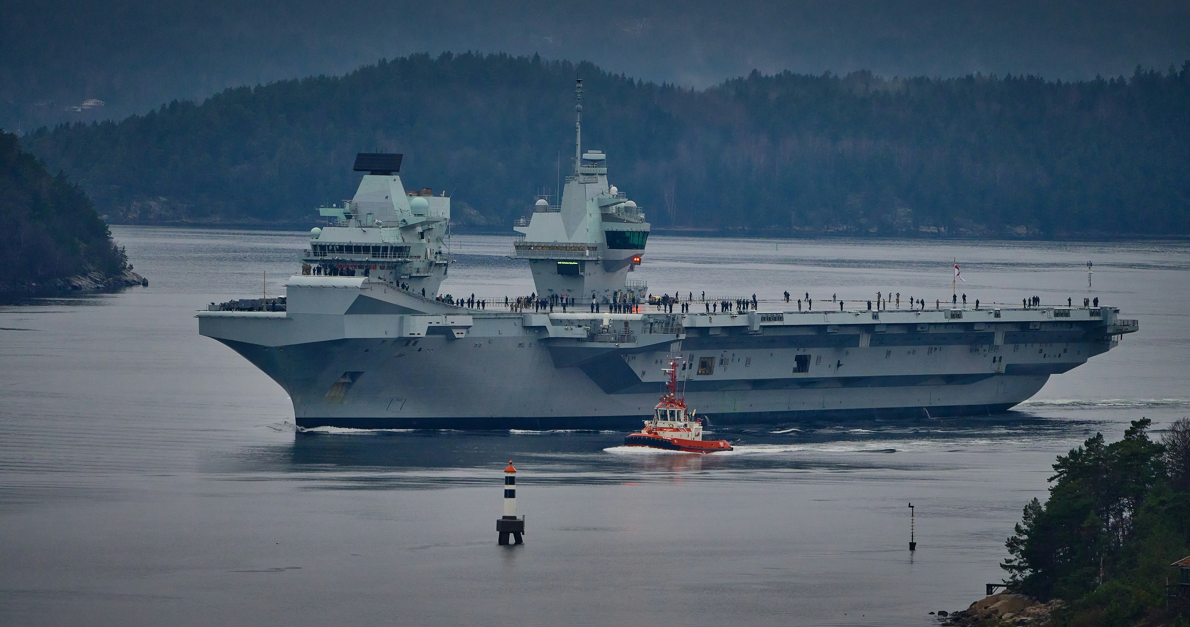 A large aircraft carrier gliding through calm waters, escorted by a tugboat, surrounded by a serene landscape of trees and distant hills.