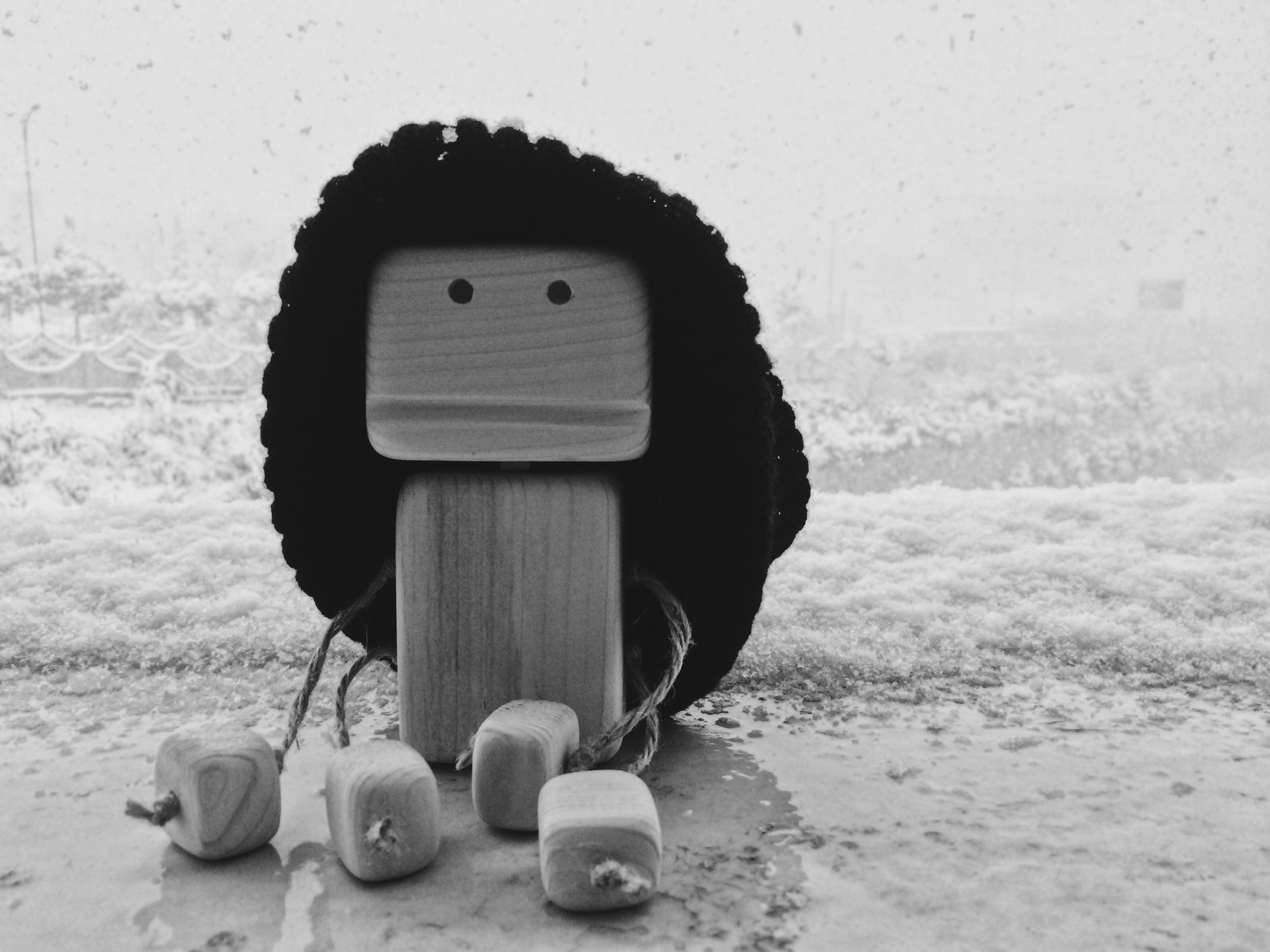 Wooden figure wearing a knitted hat stands amidst a snowy landscape, surrounded by small white blocks resembling snowballs. A sense of solitude and whimsy permeates the scene.