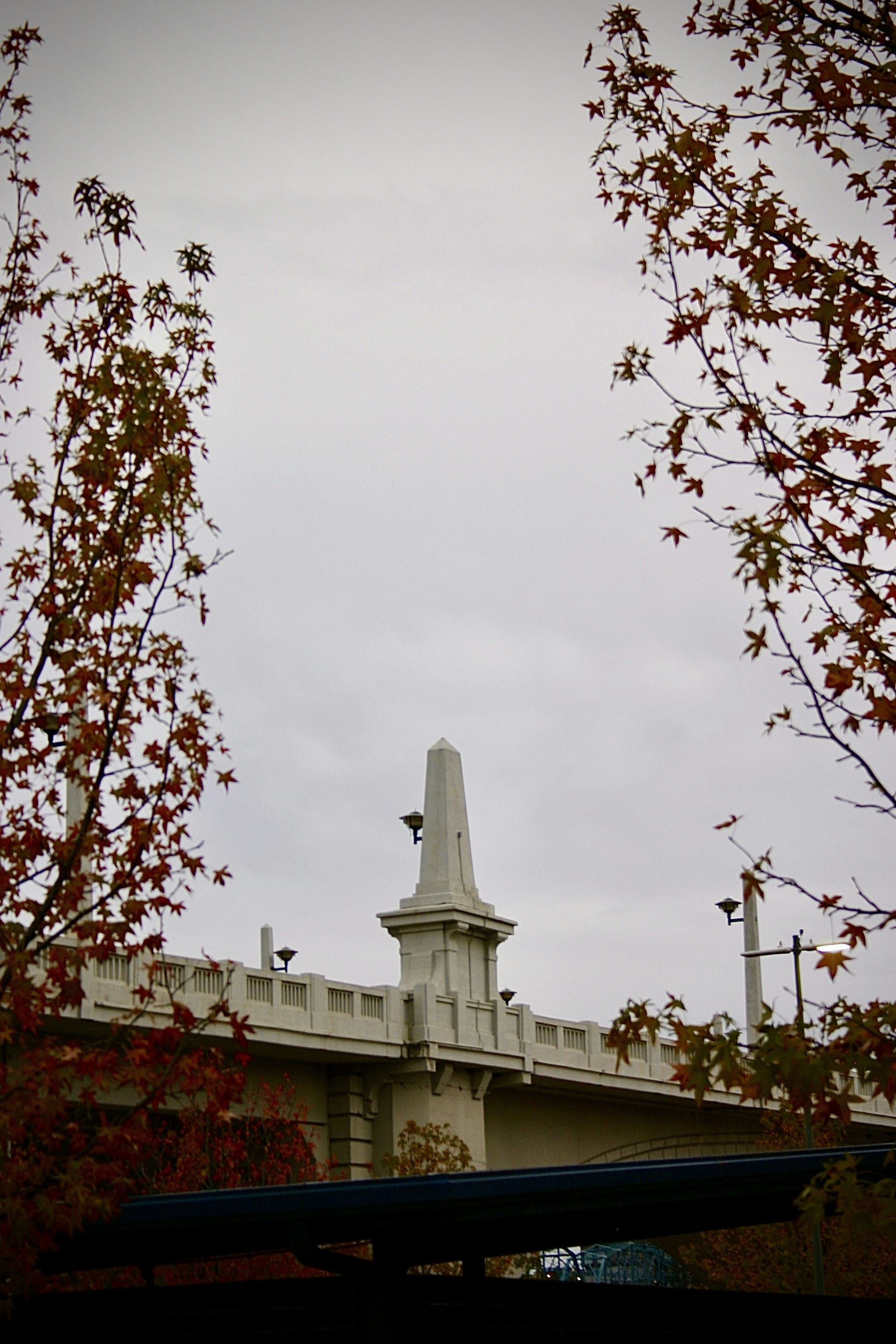Monument obelisk on a bridge framed by autumn foliage beneath a gray sky.