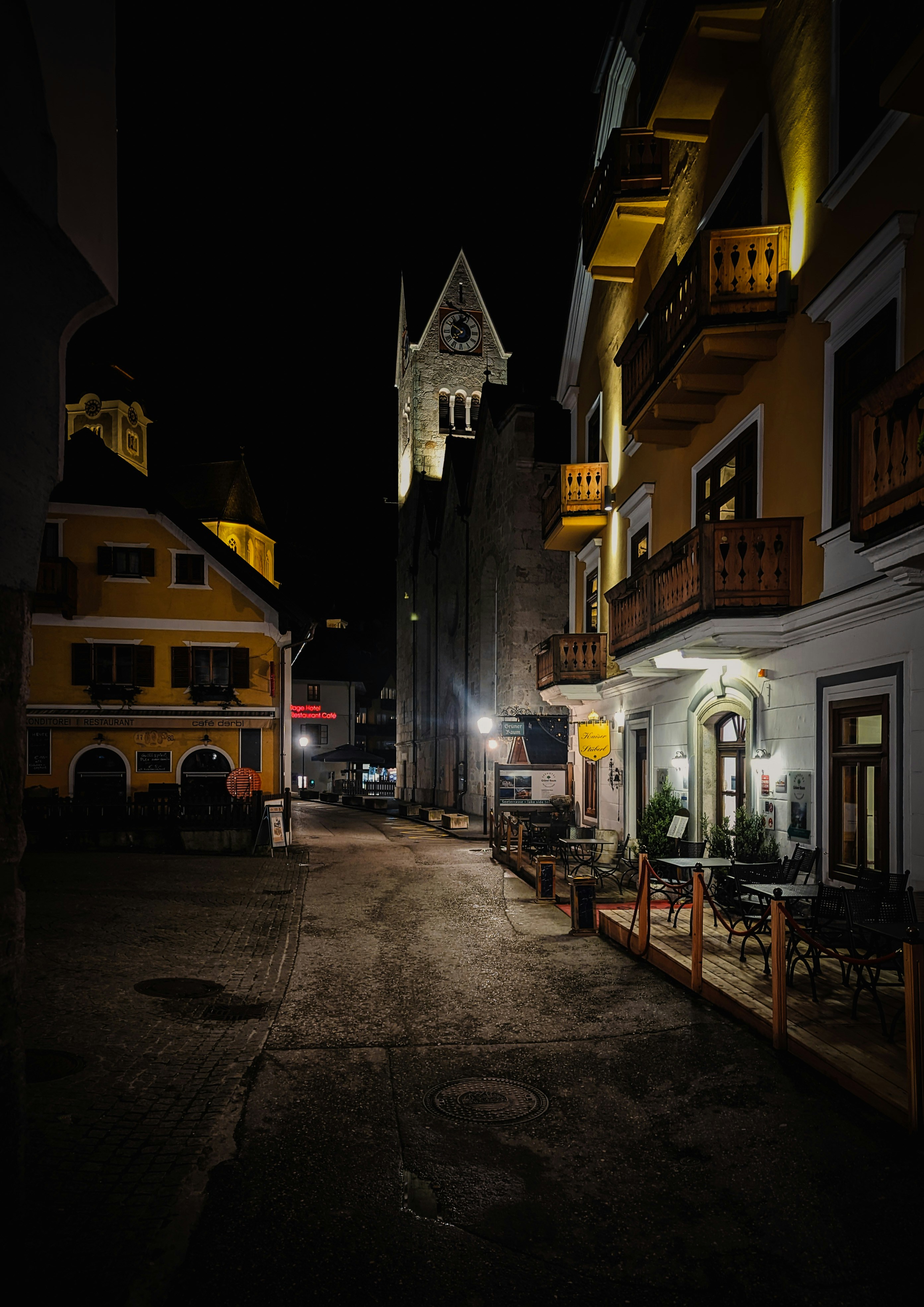 Night street scene photographed, featuring a clock tower rising above illuminated buildings. Warm lights cast on a quiet cobbled avenue with outdoor seating.