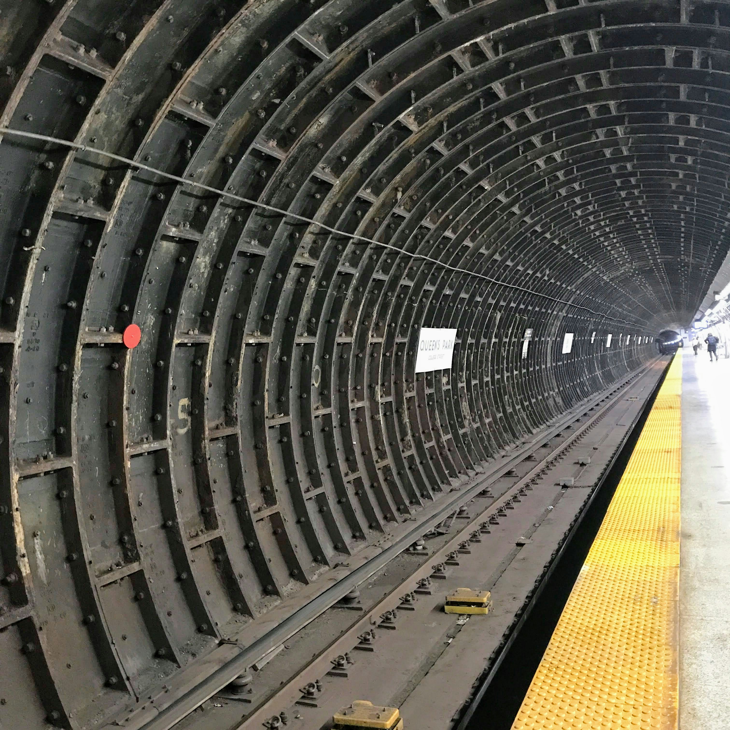 View of an underground train tunnel with arched metal walls and yellow platform edge, leading into a distant light source.