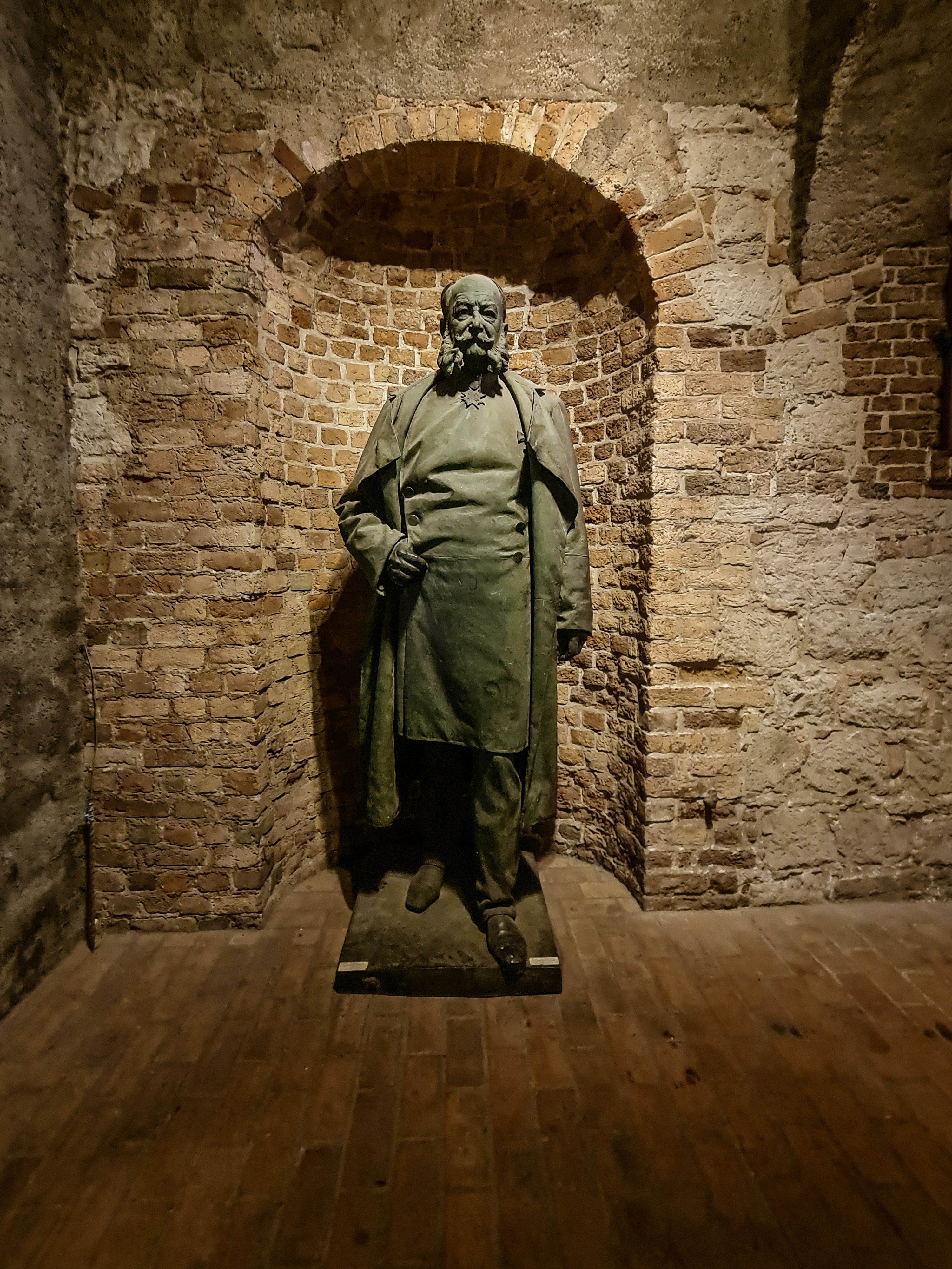Patina-green statue of a robed sage stands in a brick-arched alcove inside a dim wooden-floored chamber.
